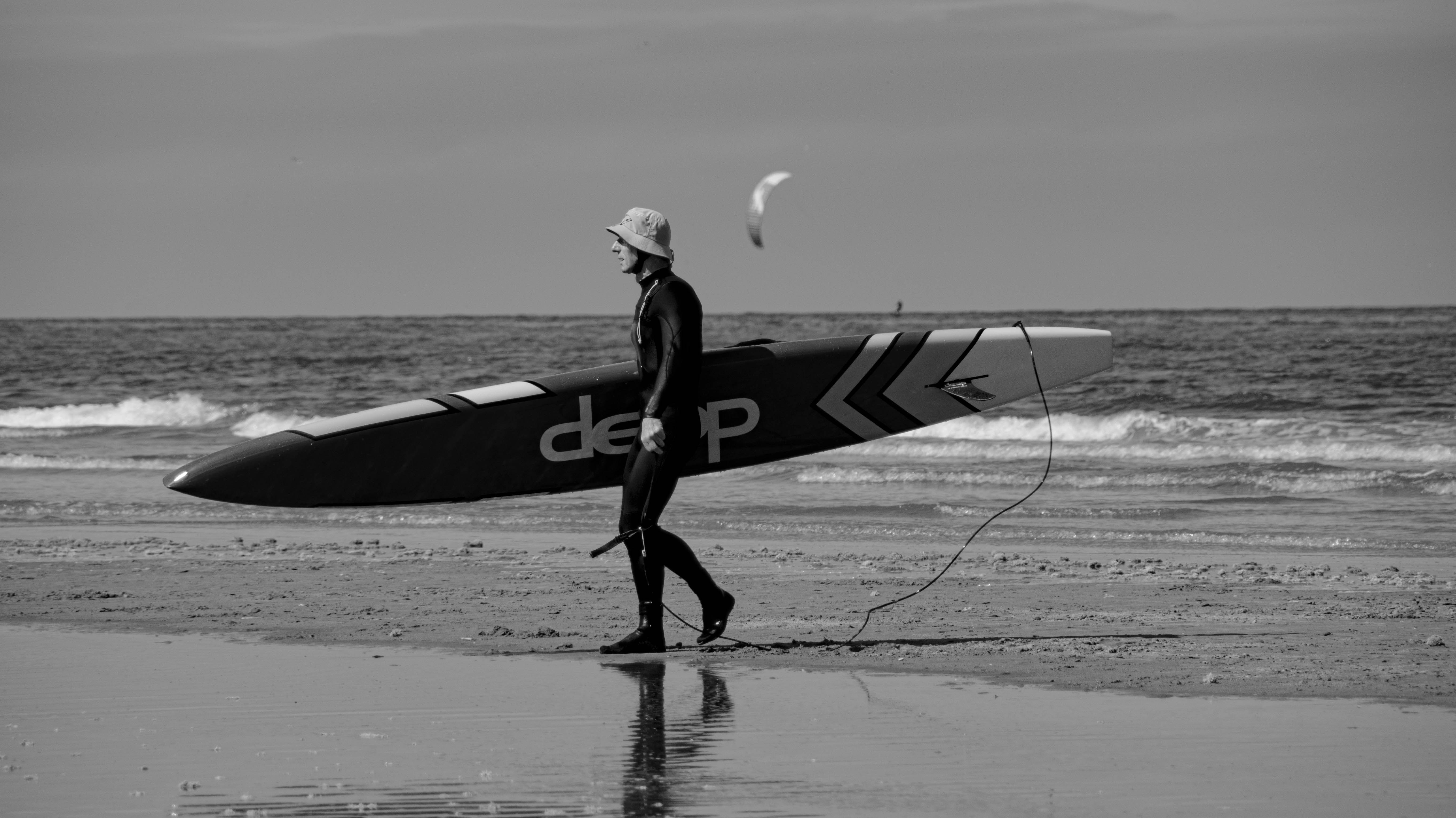 Gray Tidewalk: A lone surfer drags a weathered board along a reflective shoreline
