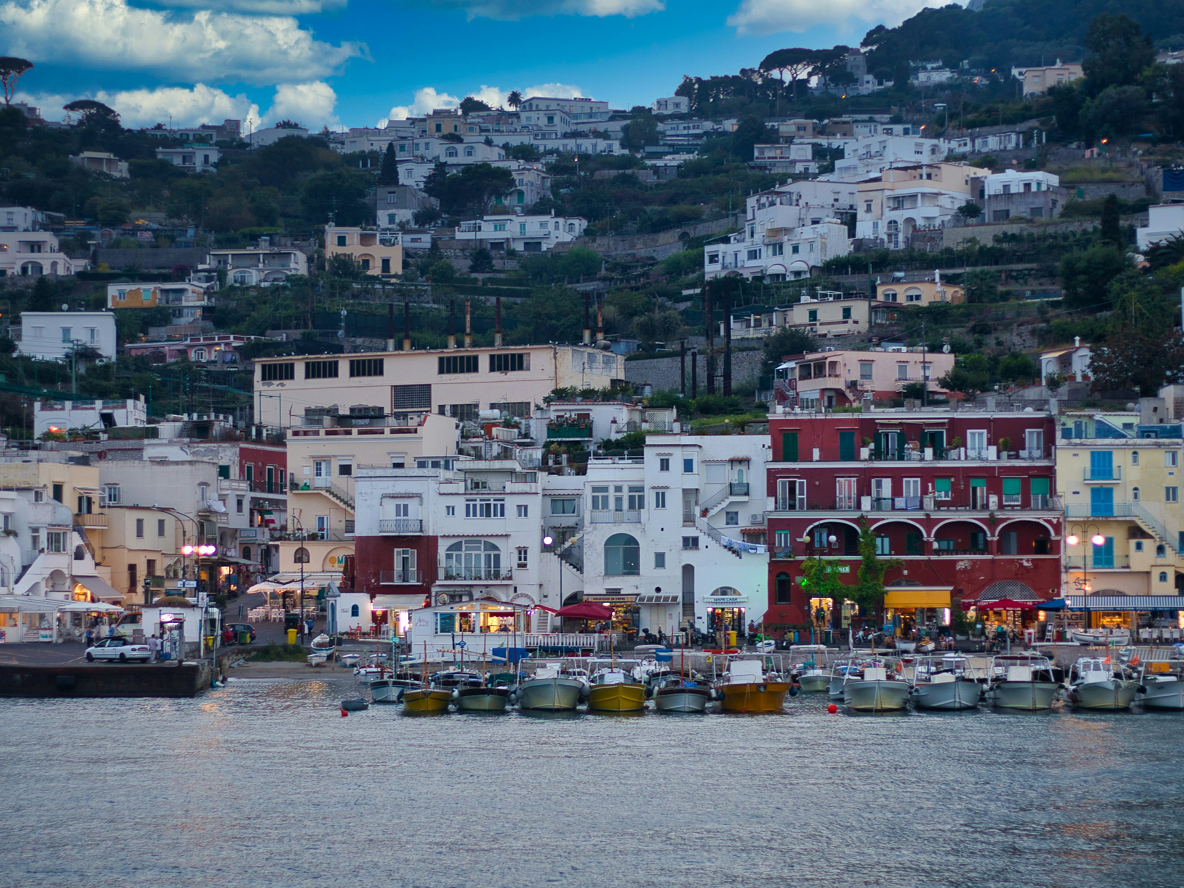 Charming coastal village with colorful buildings lining the harbor, illuminated by warm lights as dusk settles. Fishing boats rest peacefully in the water.