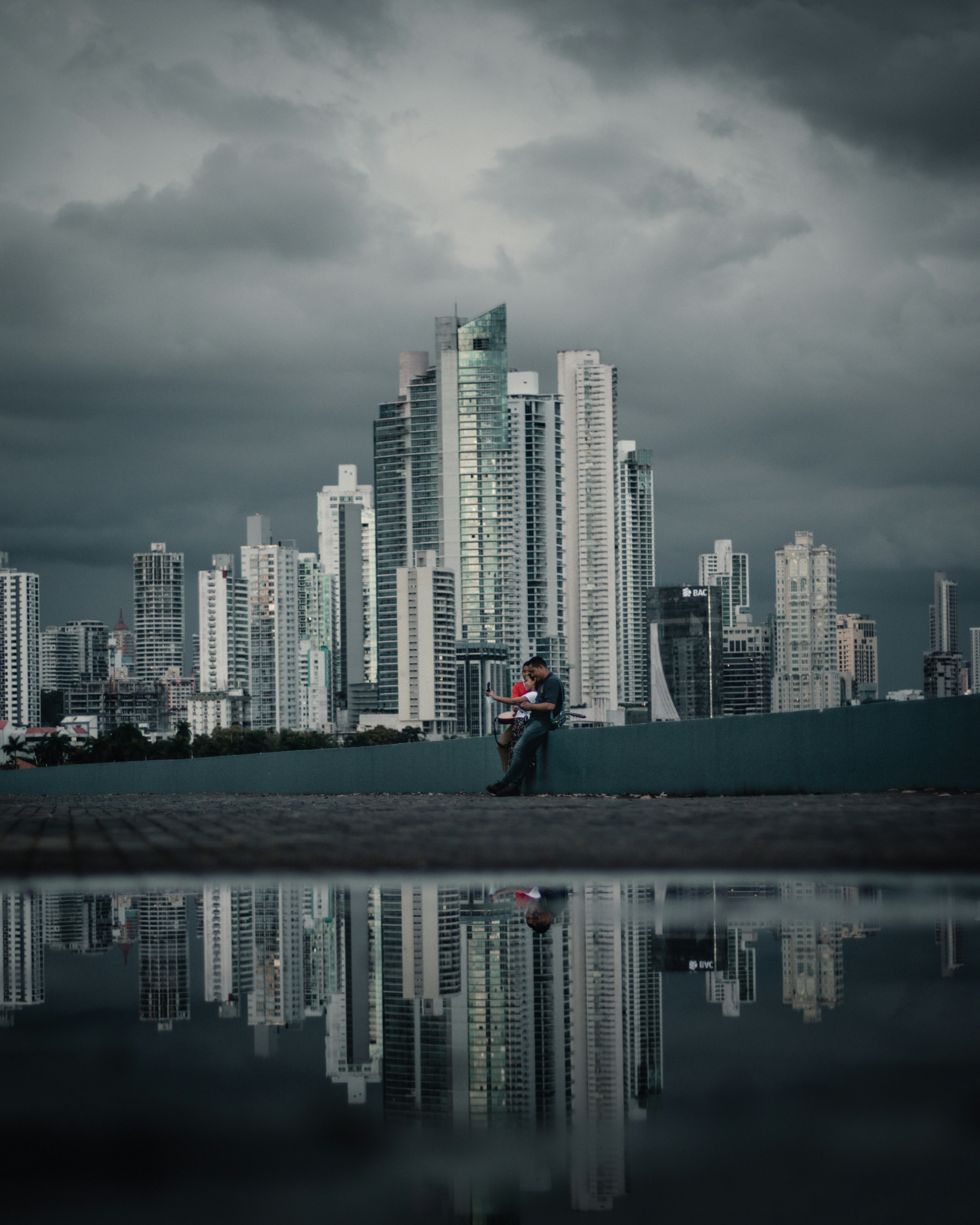 A lone figure sitting on a ledge, engrossed in a device, with towering skyscrapers and a moody sky reflected in a puddle nearby.