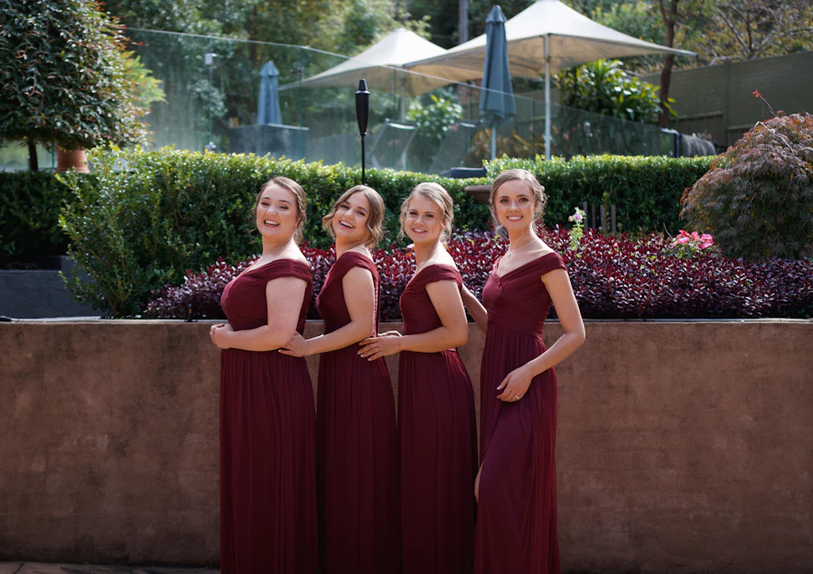 Four women in matching burgundy off-shoulder dresses stand together outdoors, smiling in front of manicured hedges and patio umbrellas. Bright daylight and layered garden greenery frame the group portrait.