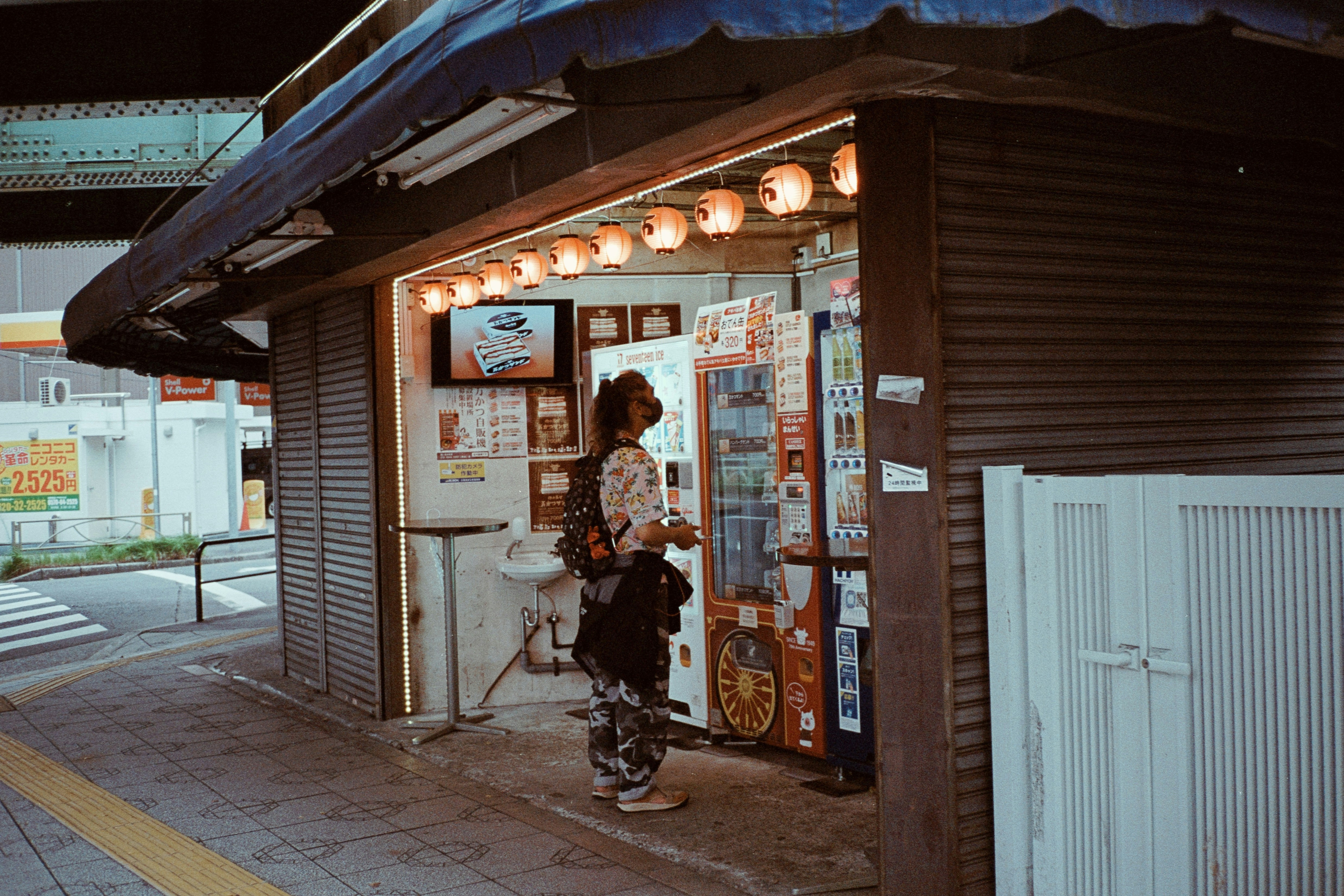 A woman browses a vending display outside a lantern-lit street stand at dusk.