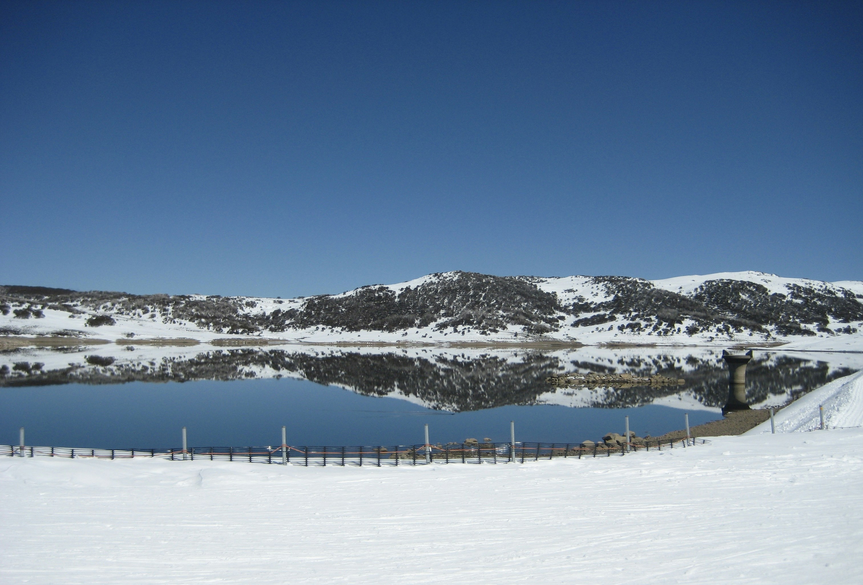 Snow-covered landscape with a calm lake reflecting the ridge and a weathered fence along the shore. A small structure sits by the water's edge under a clear blue sky.