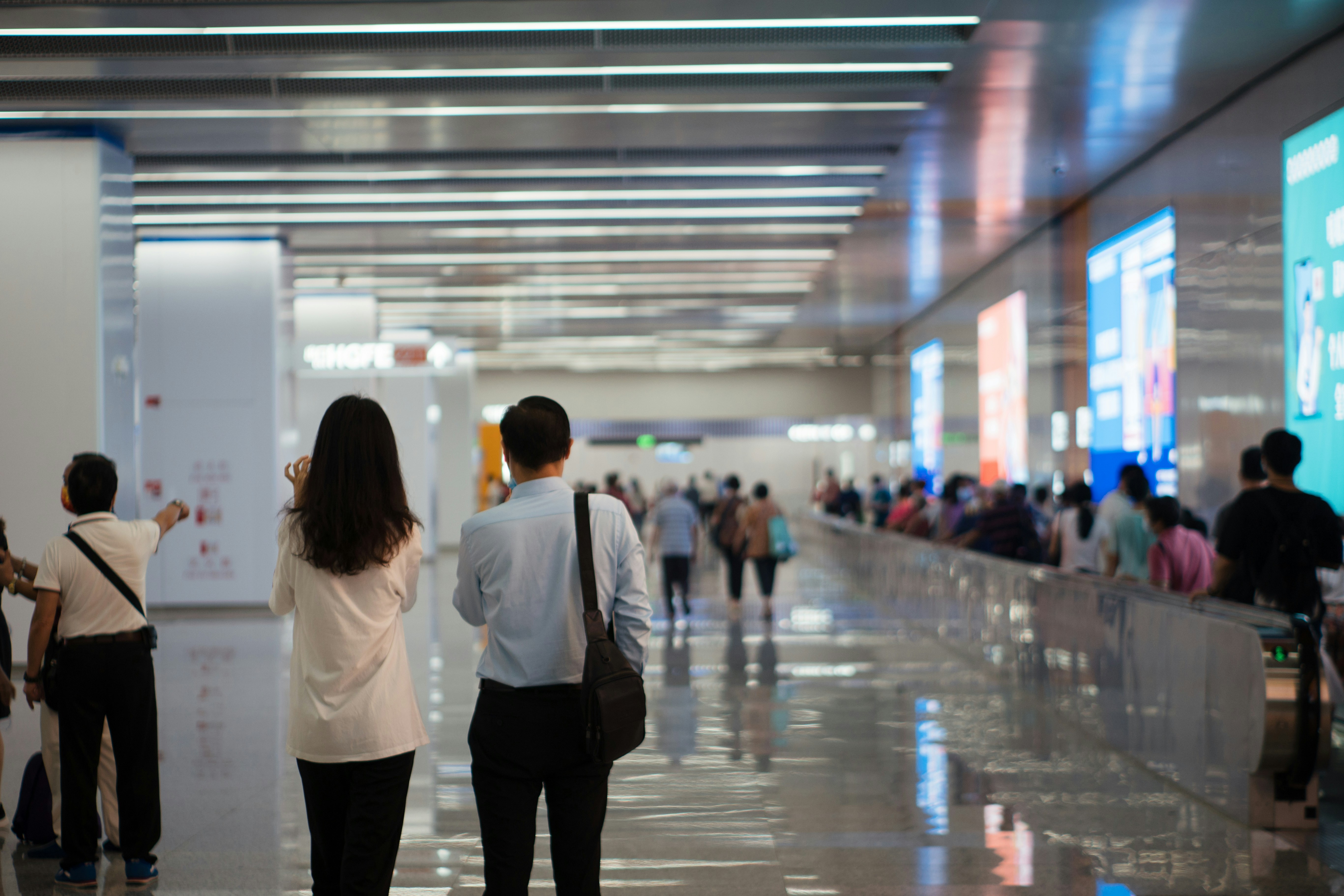 Two individuals walking in a bustling terminal, surrounded by a diverse crowd and illuminated advertising displays.