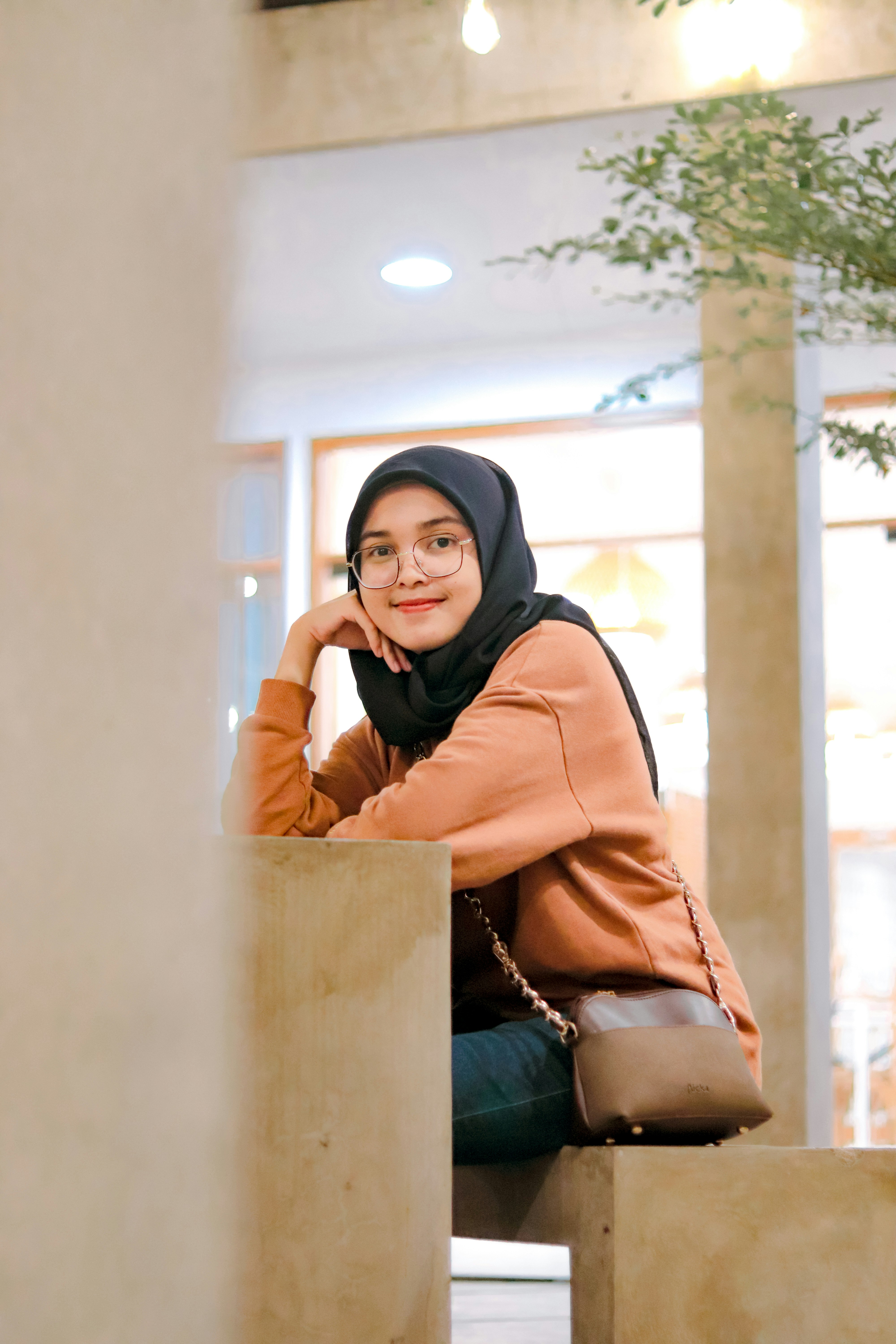 Woman in a hijab sitting on concrete steps, smiling in a softly lit modern interior.