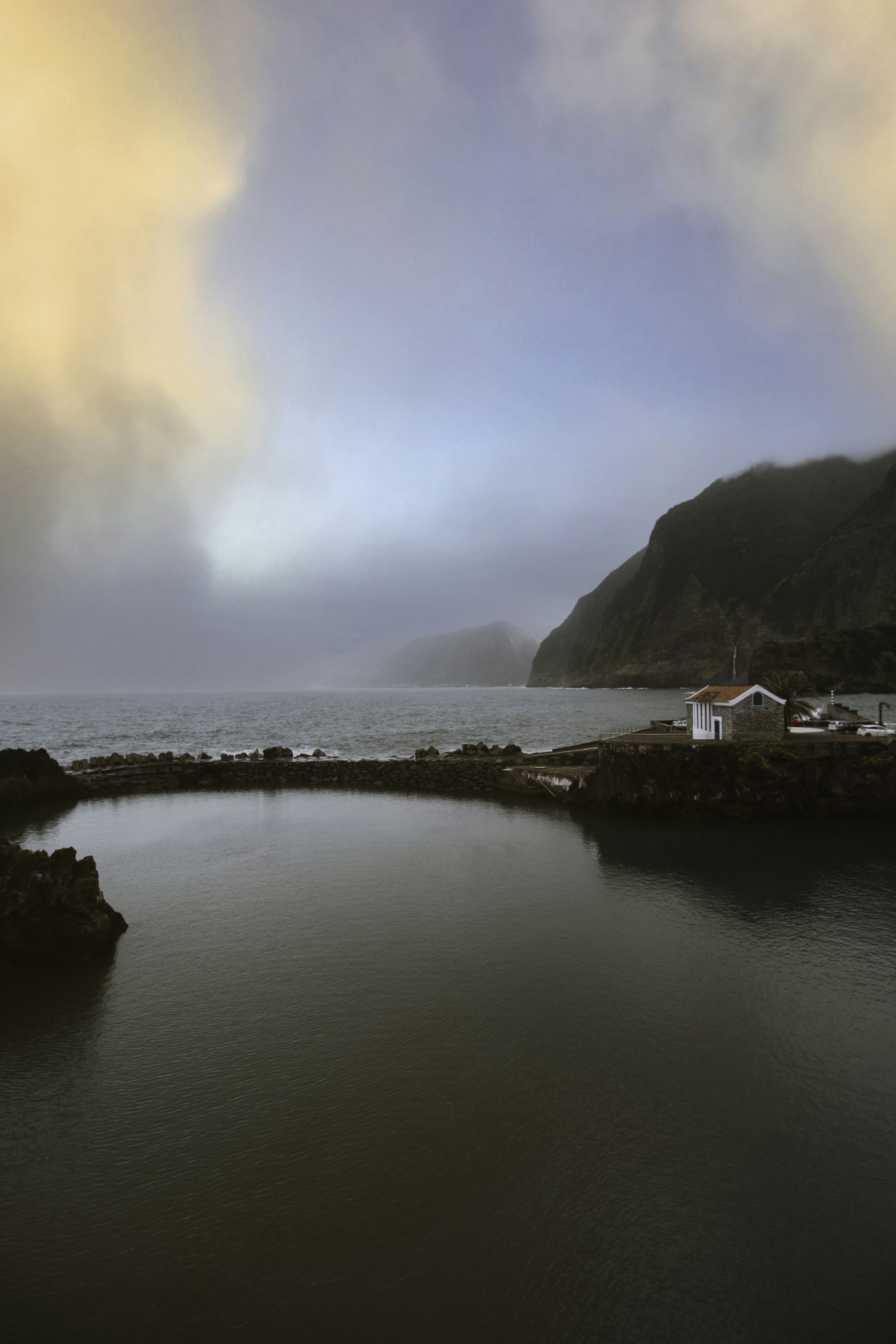 Lone white house by a tranquil harbor, shrouded in misty coastal fog with rugged cliffs in the background.