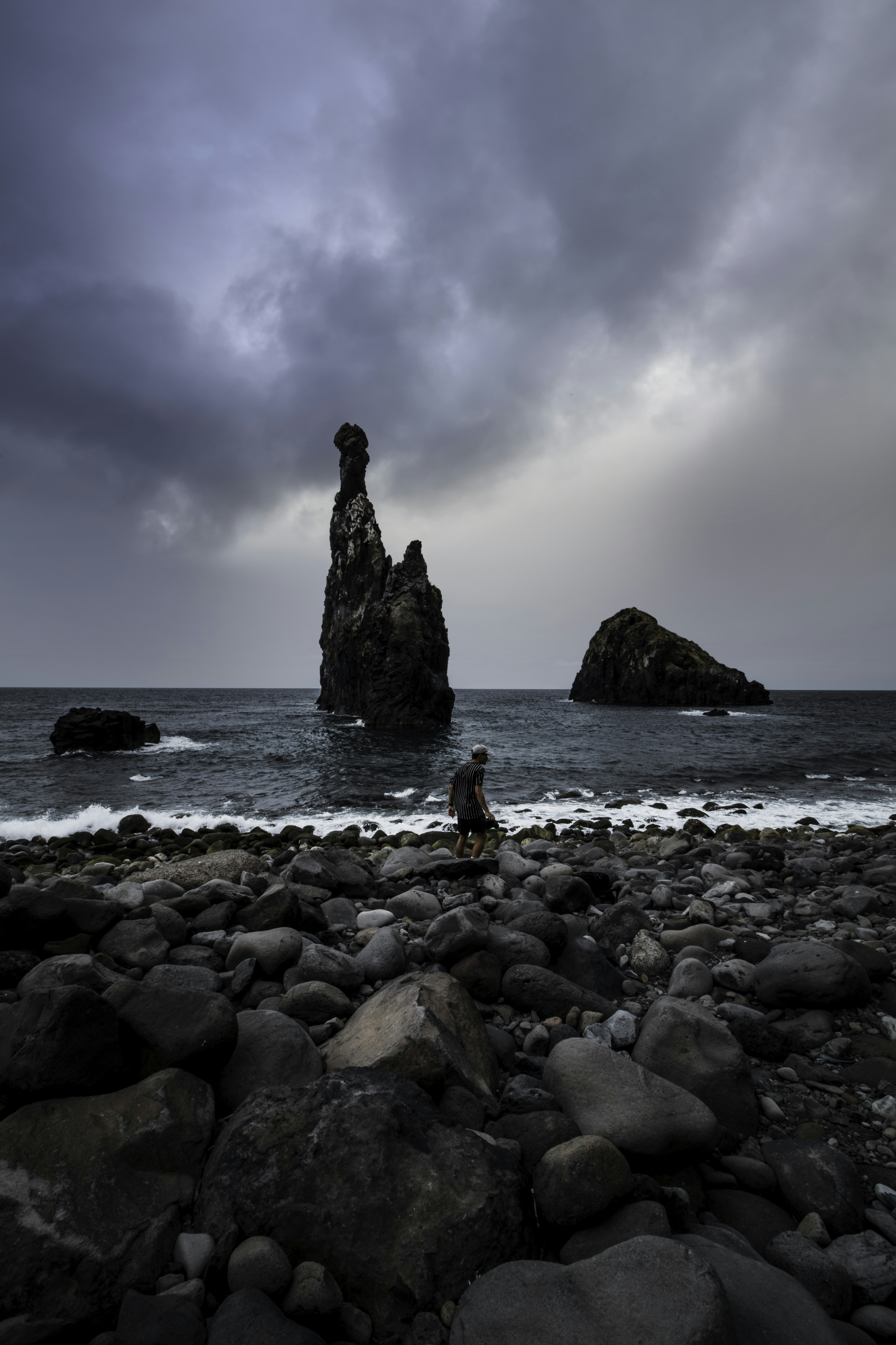 A lone figure walks along a rocky beach, gazing at towering sea stacks under a moody sky. The waves crash gently against the stones.