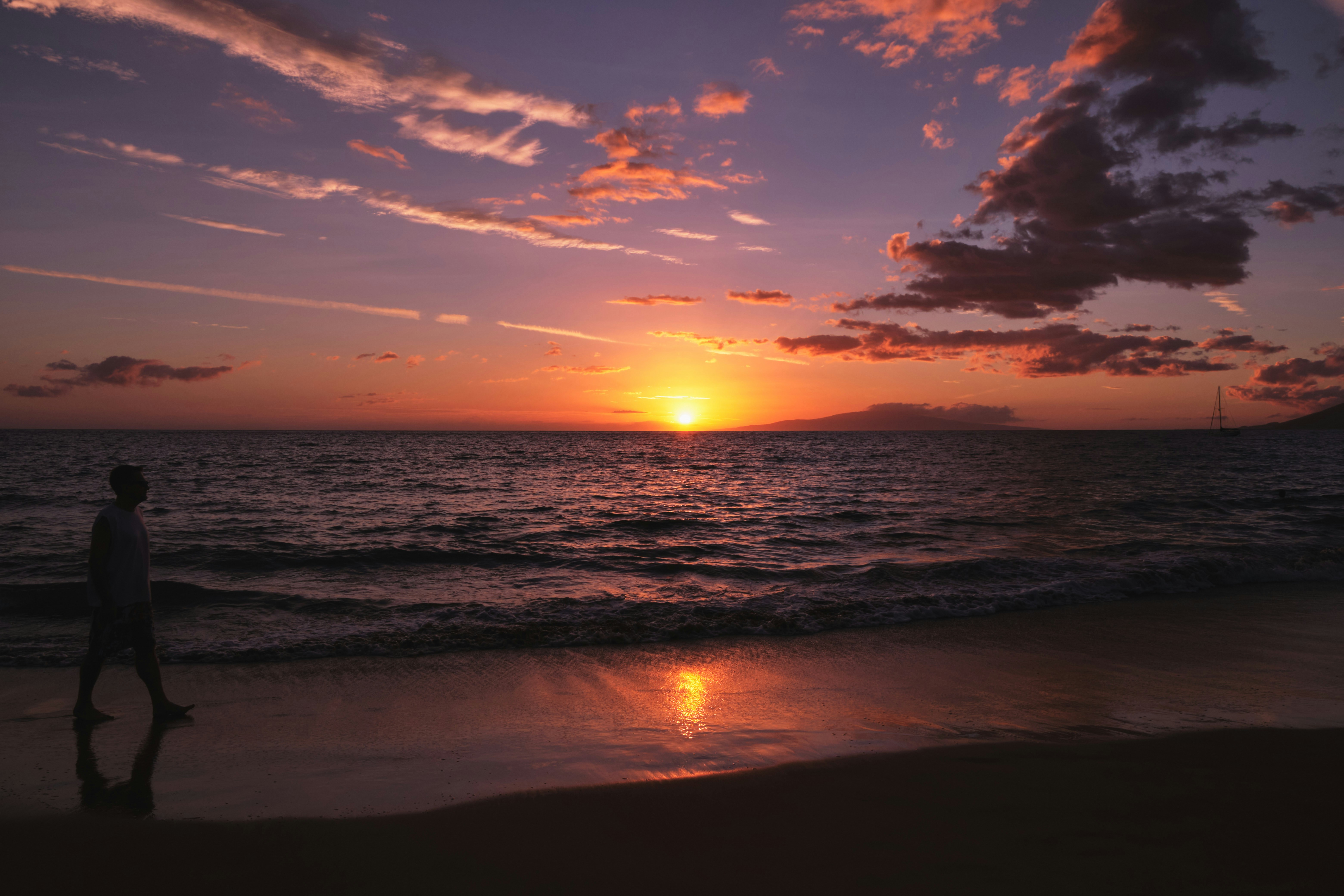 Silhouette of a person walking along the shoreline as the sun sets over the horizon, casting vibrant colors across the sky and reflecting on the water.