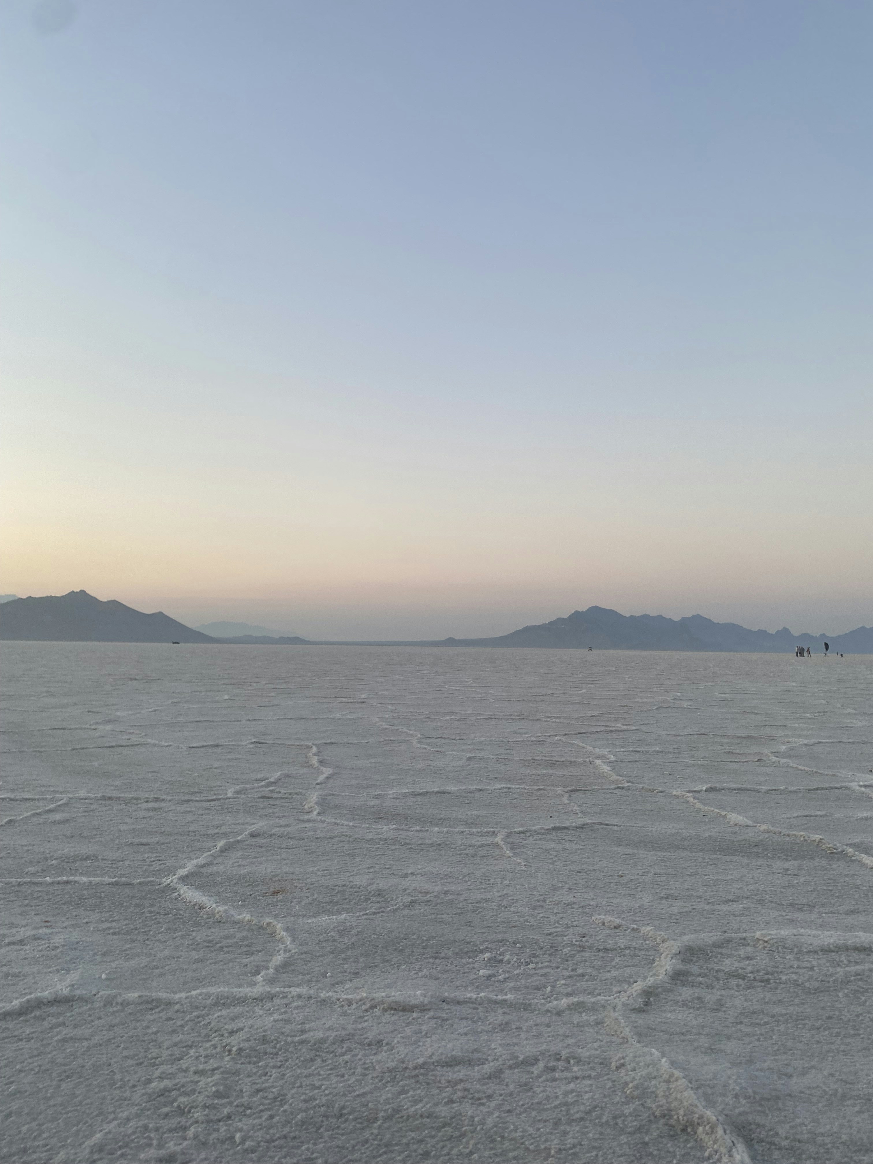 Expansive salt flats stretch towards distant mountains under a soft twilight sky, with tiny figures walking along the horizon.