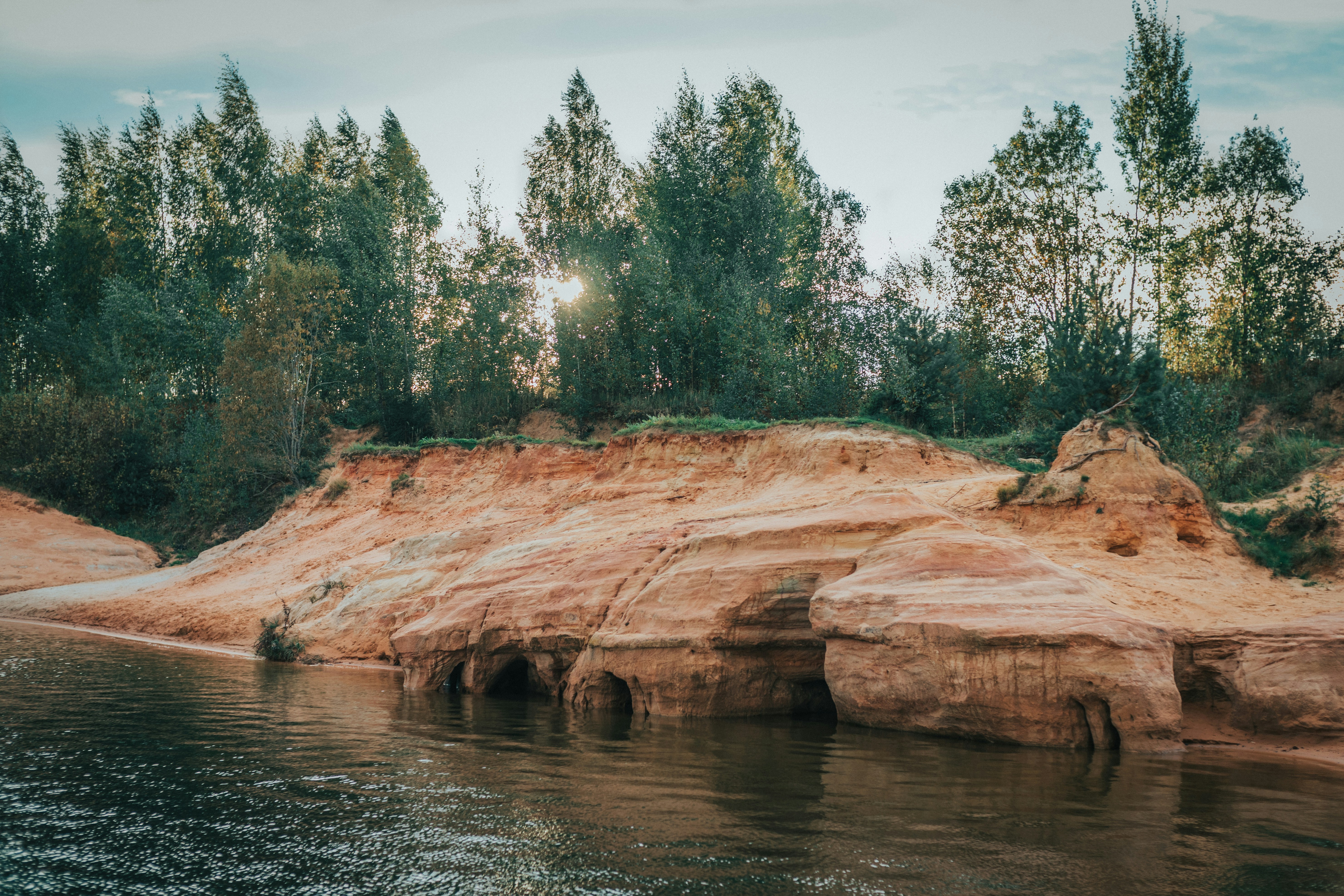 Sandy cliffs along the quarry lake near Novinka, Leningrad Region. The warm-toned sandstone formations with small natural arches descend into the calm water, framed by birch and pine trees under the soft evening light.
