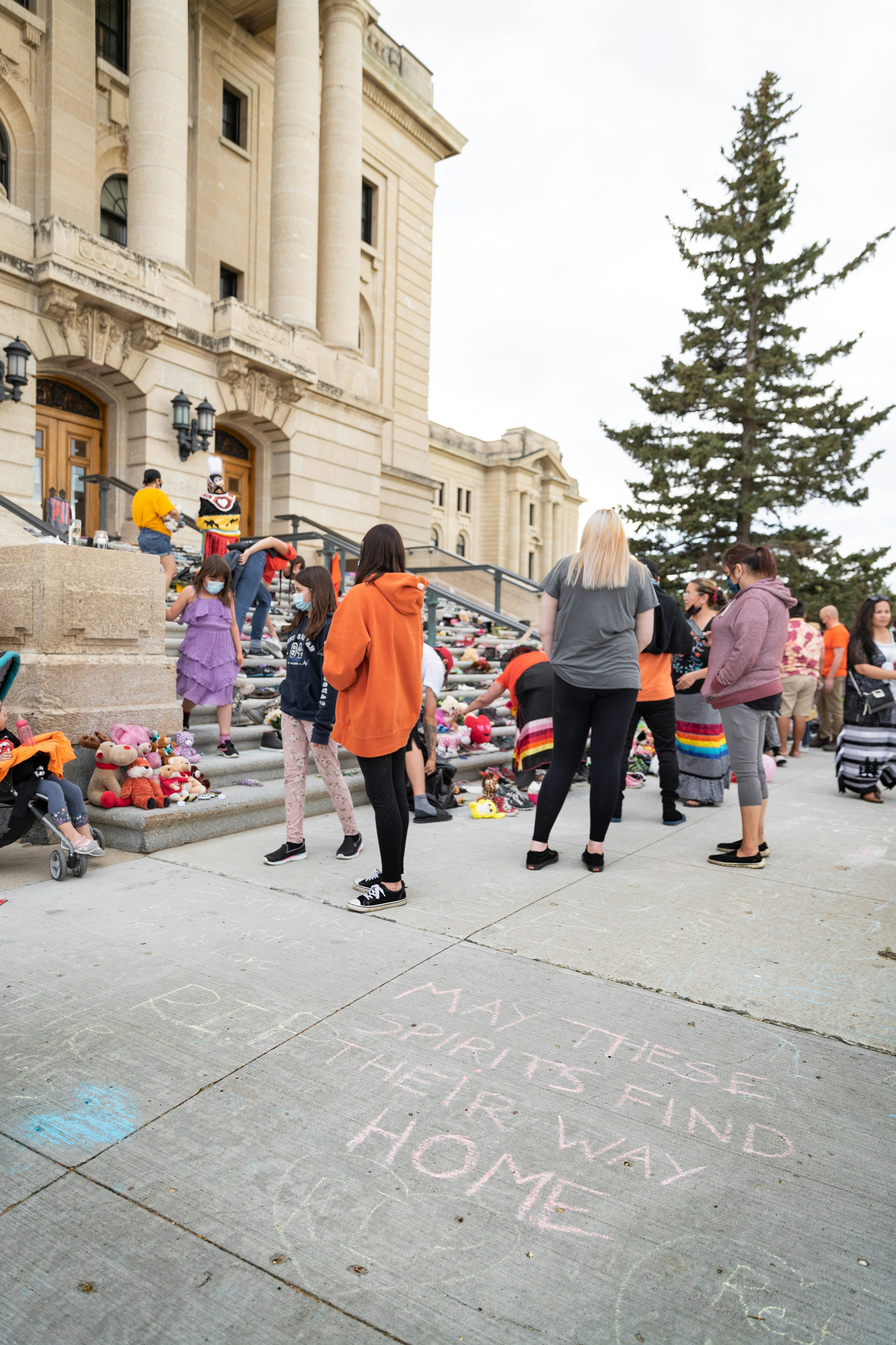 Community members gather on the steps of a historic building, surrounded by memorial items and messages written in chalk. A poignant tribute to lost spirits and the importance of remembrance.