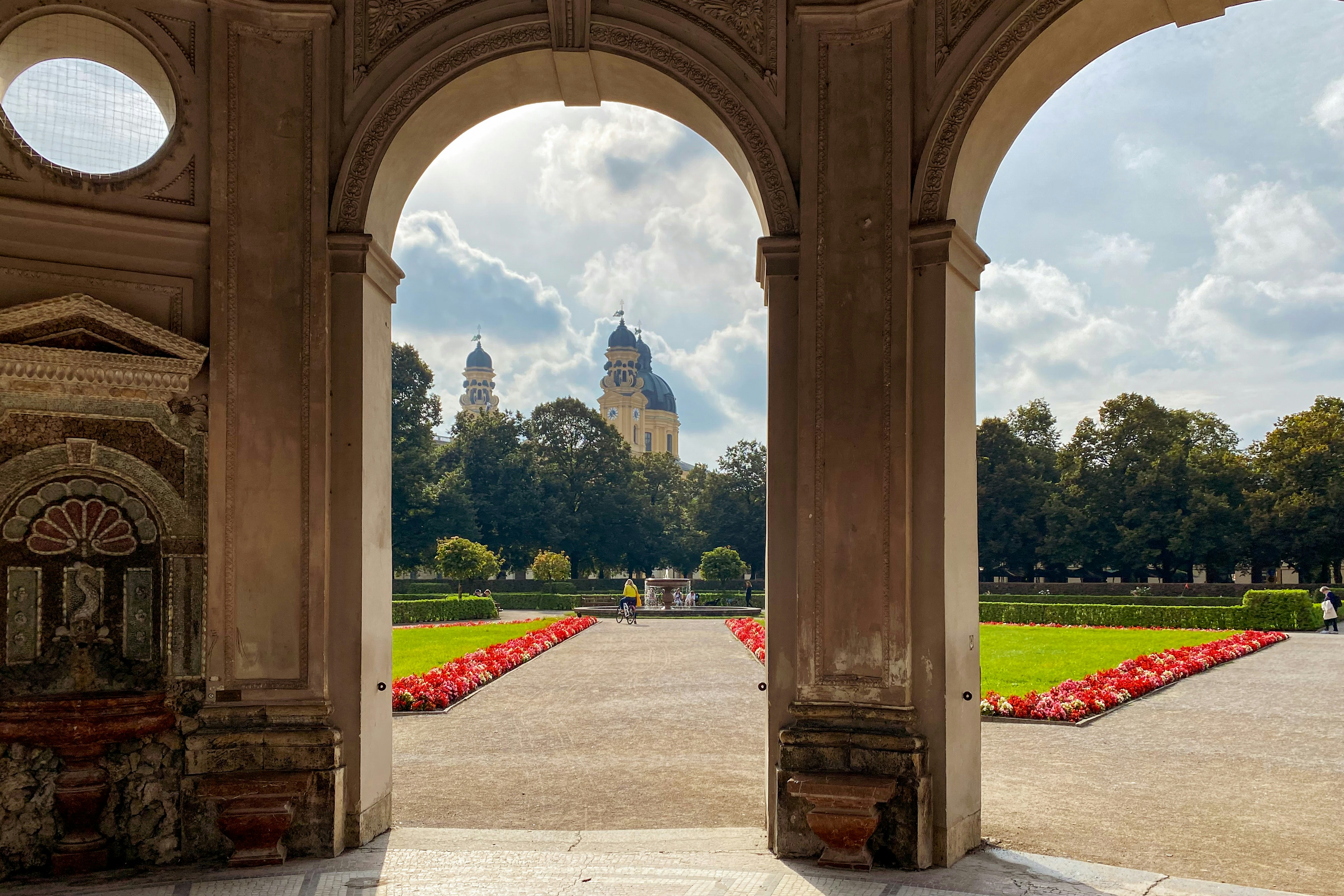 View through ornate arches leading to a vibrant garden with blooming flowers and distant architectural details. The scene invites tranquility and exploration.