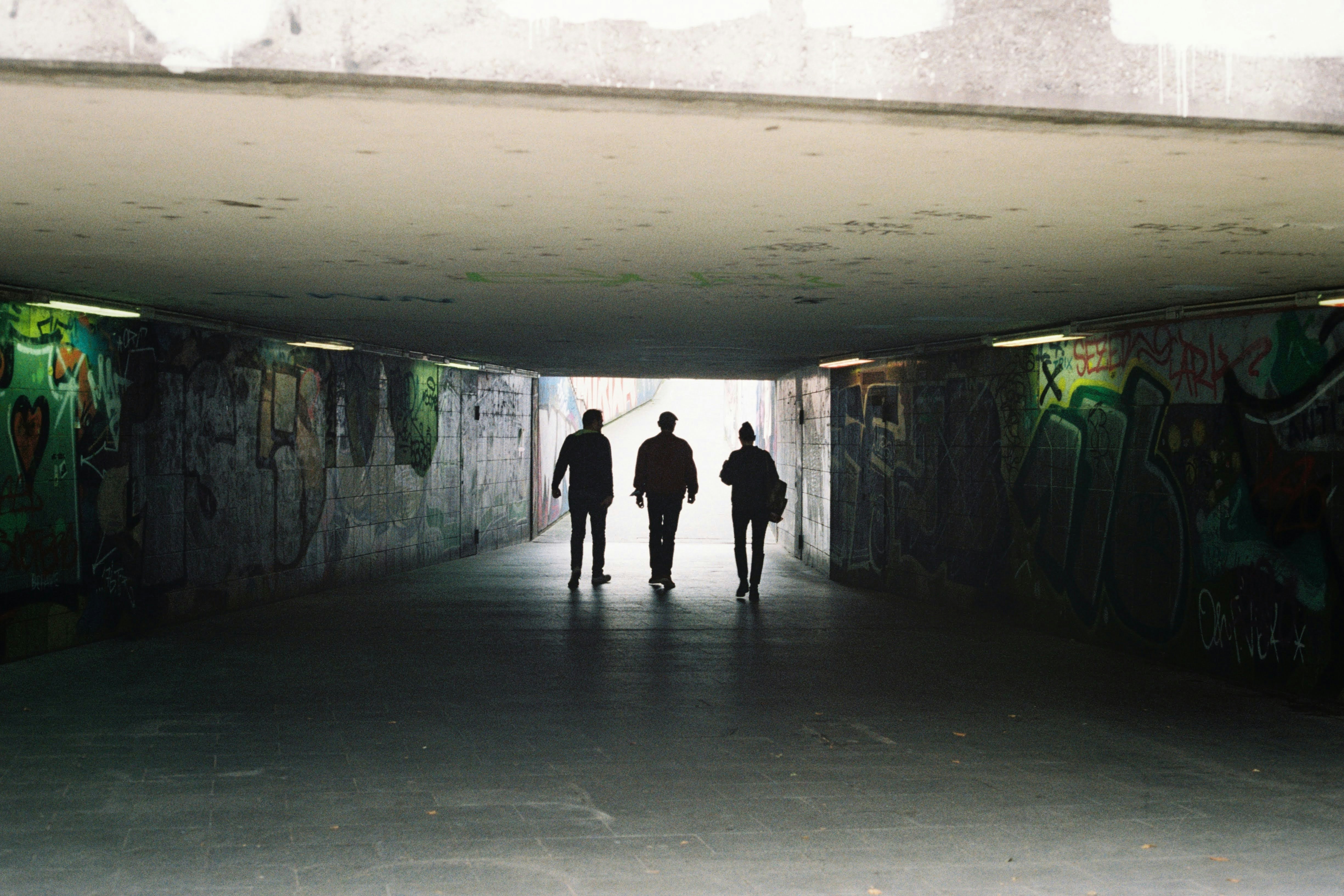 Three silhouetted figures walk toward a bright exit in a graffiti-covered underpass, creating a stark backlit scene.