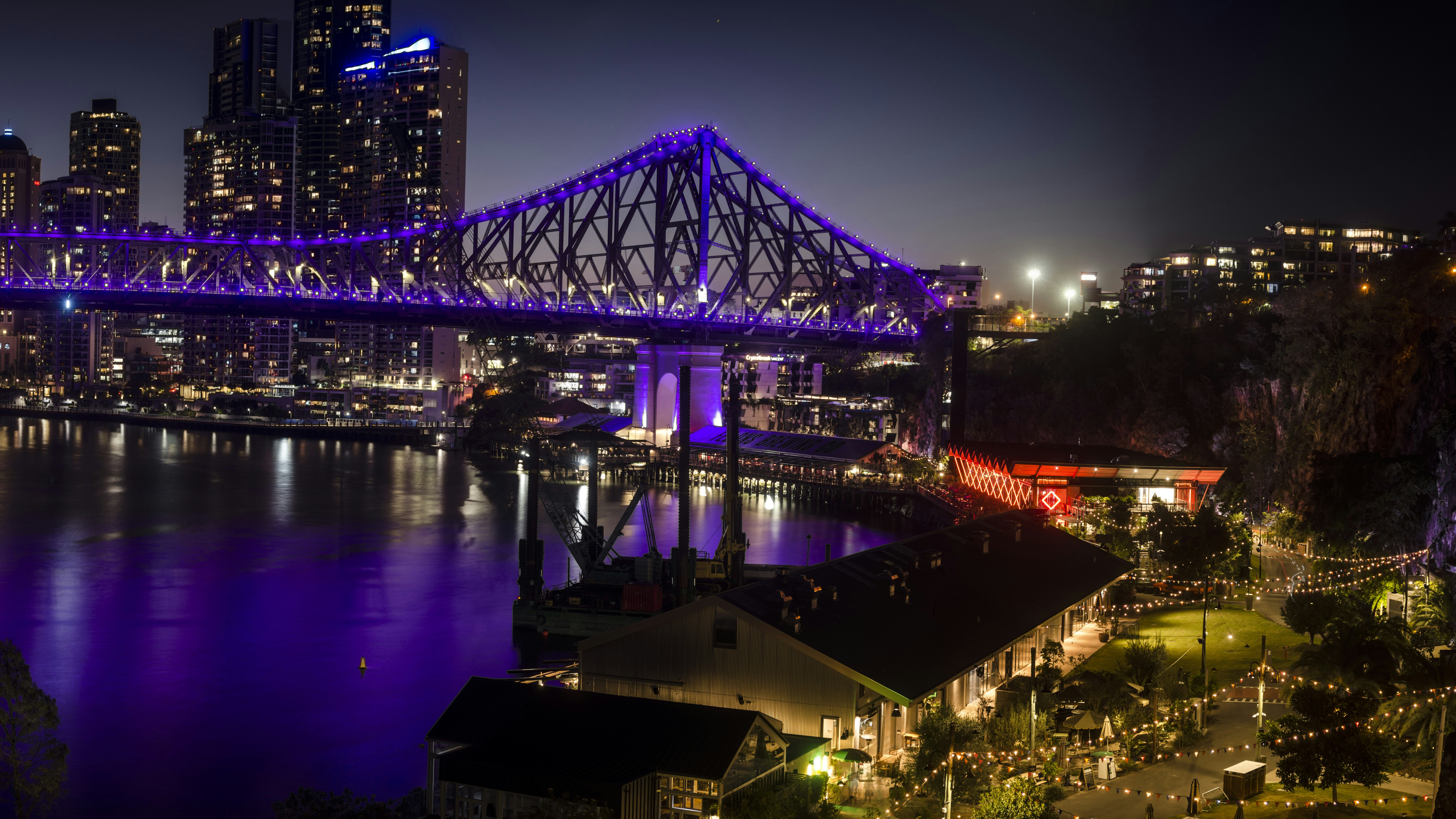 Vibrant cityscape featuring a illuminated bridge over a river, with reflections shimmering in the water and buildings adorned with lights.