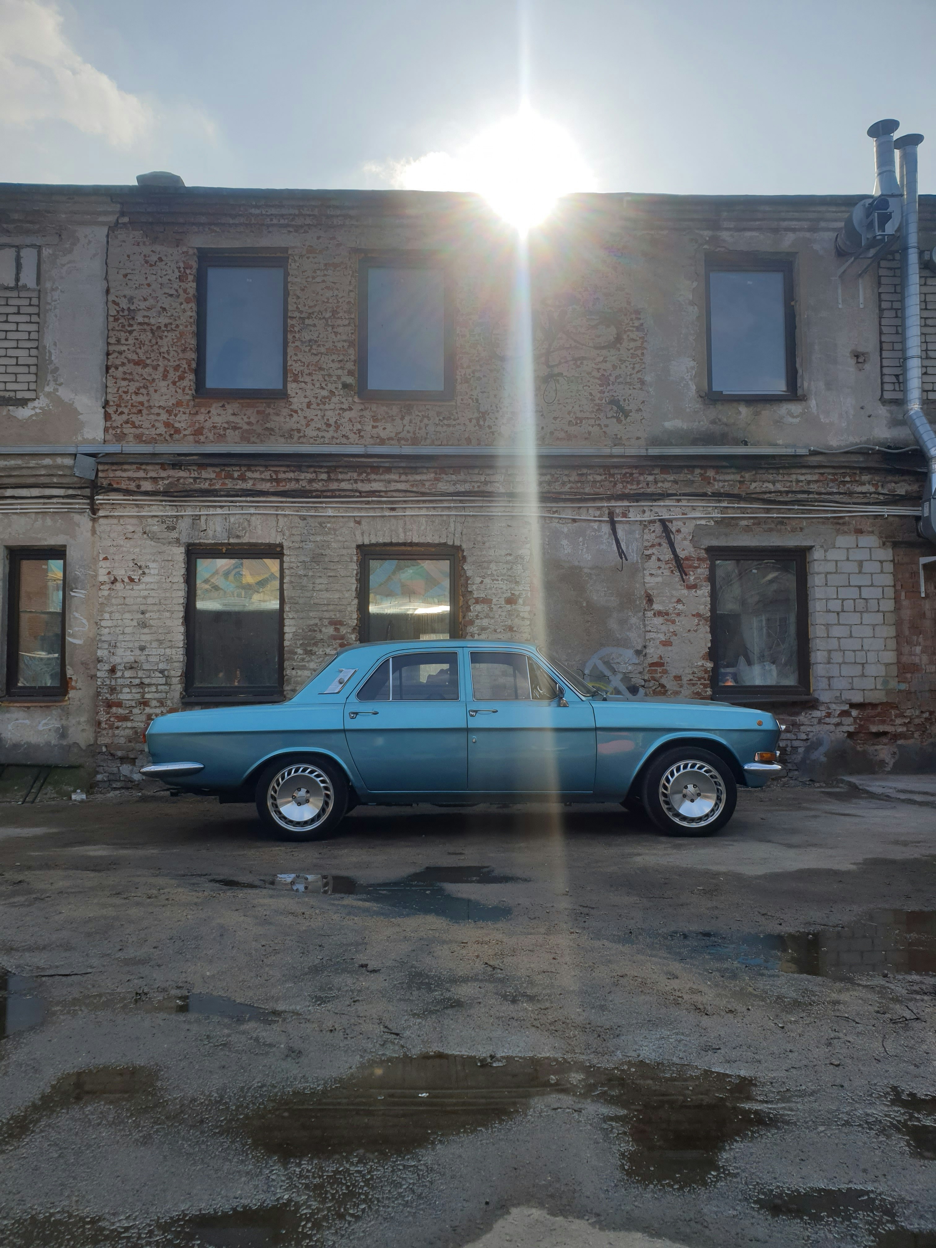 Sunlit Blue Classic: A 1960s sedan rests before a weathered brick facade