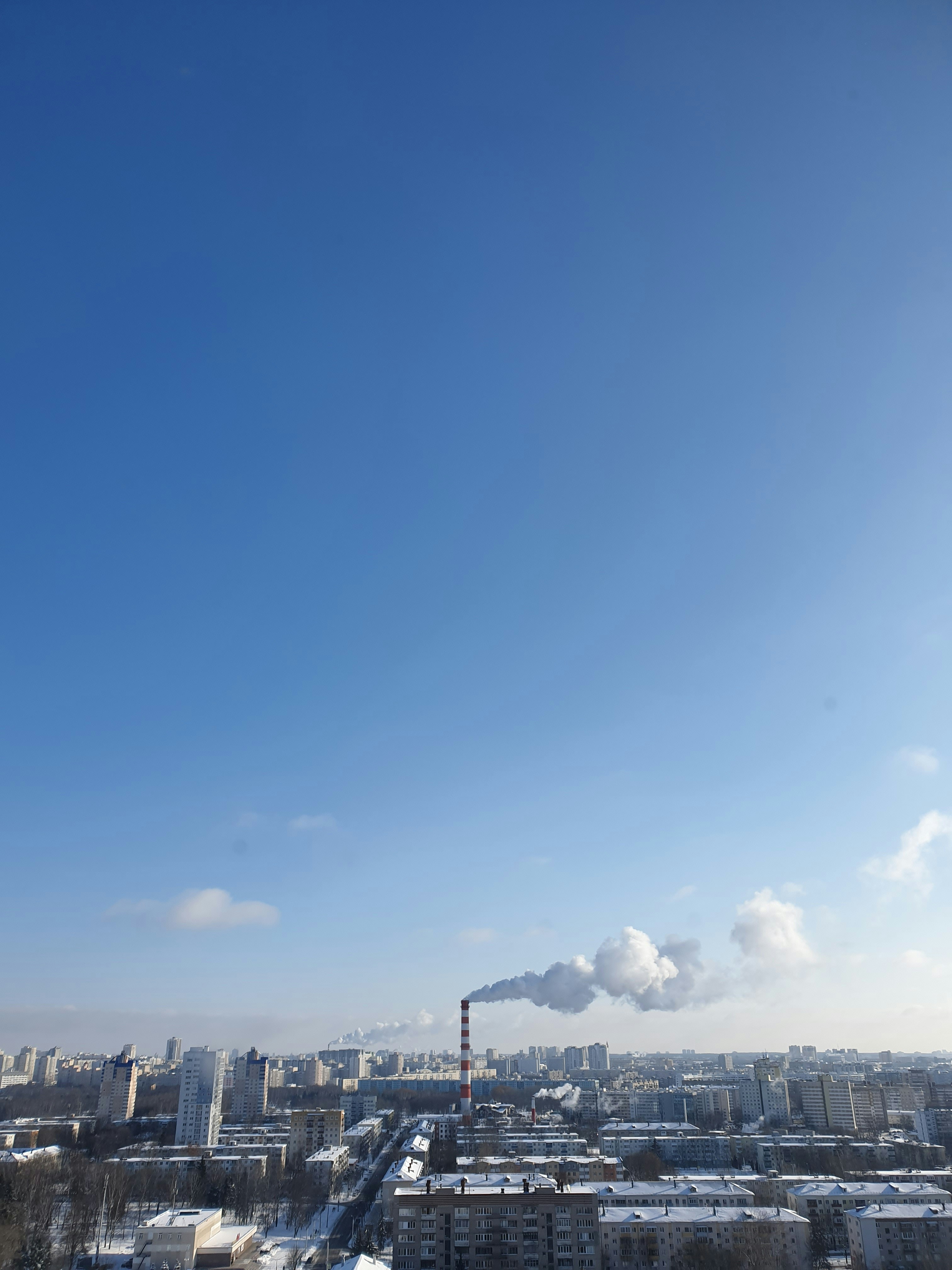 A panoramic view of a snowy cityscape under a clear blue sky, with smoke rising from industrial chimneys in the distance.