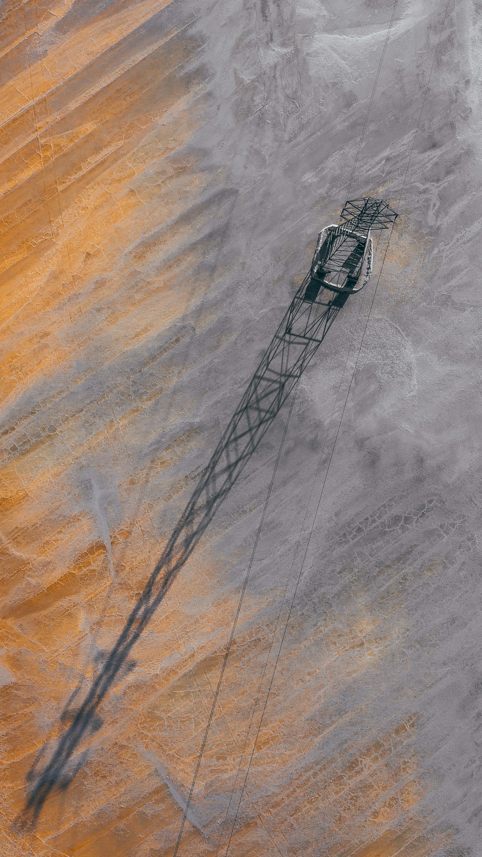 A power transmission tower casts a long shadow over a textured landscape, showcasing the interplay of light and form. The contrast between the tower and the surrounding terrain creates a striking visual narrative.