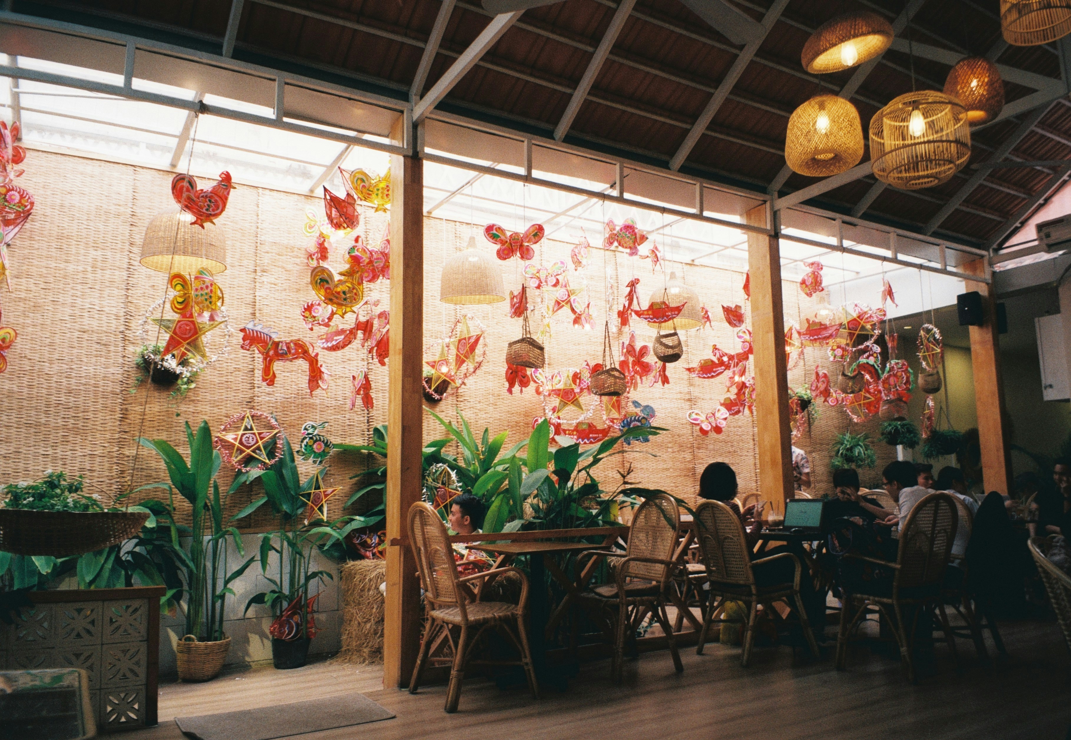 Colorful paper decorations hang from the walls of a cafe, surrounded by greenery and rustic furniture. A warm, inviting atmosphere invites guests to relax.