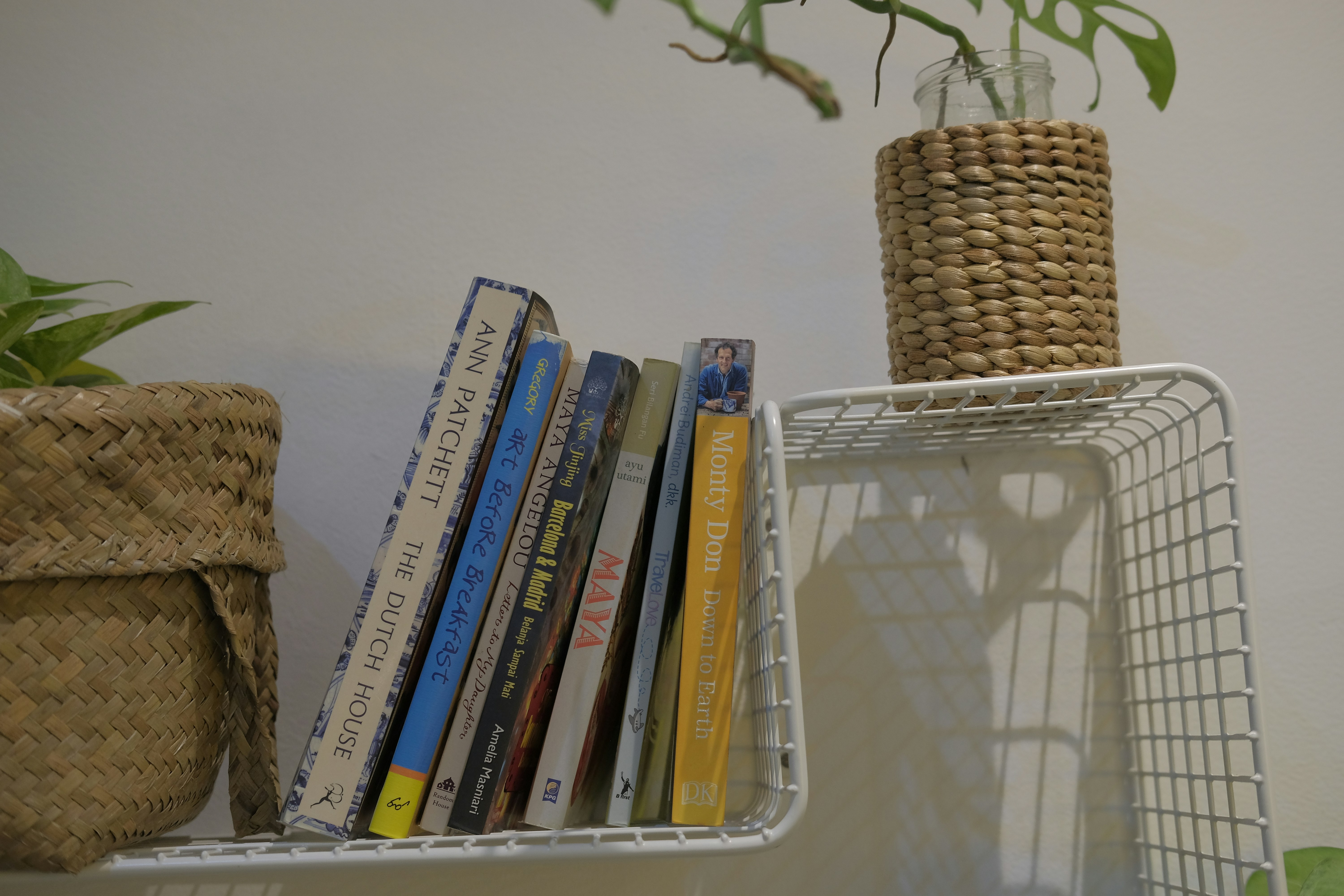 Books arranged neatly on a wire shelf beside a woven basket and a potted plant, creating a warm and inviting atmosphere.