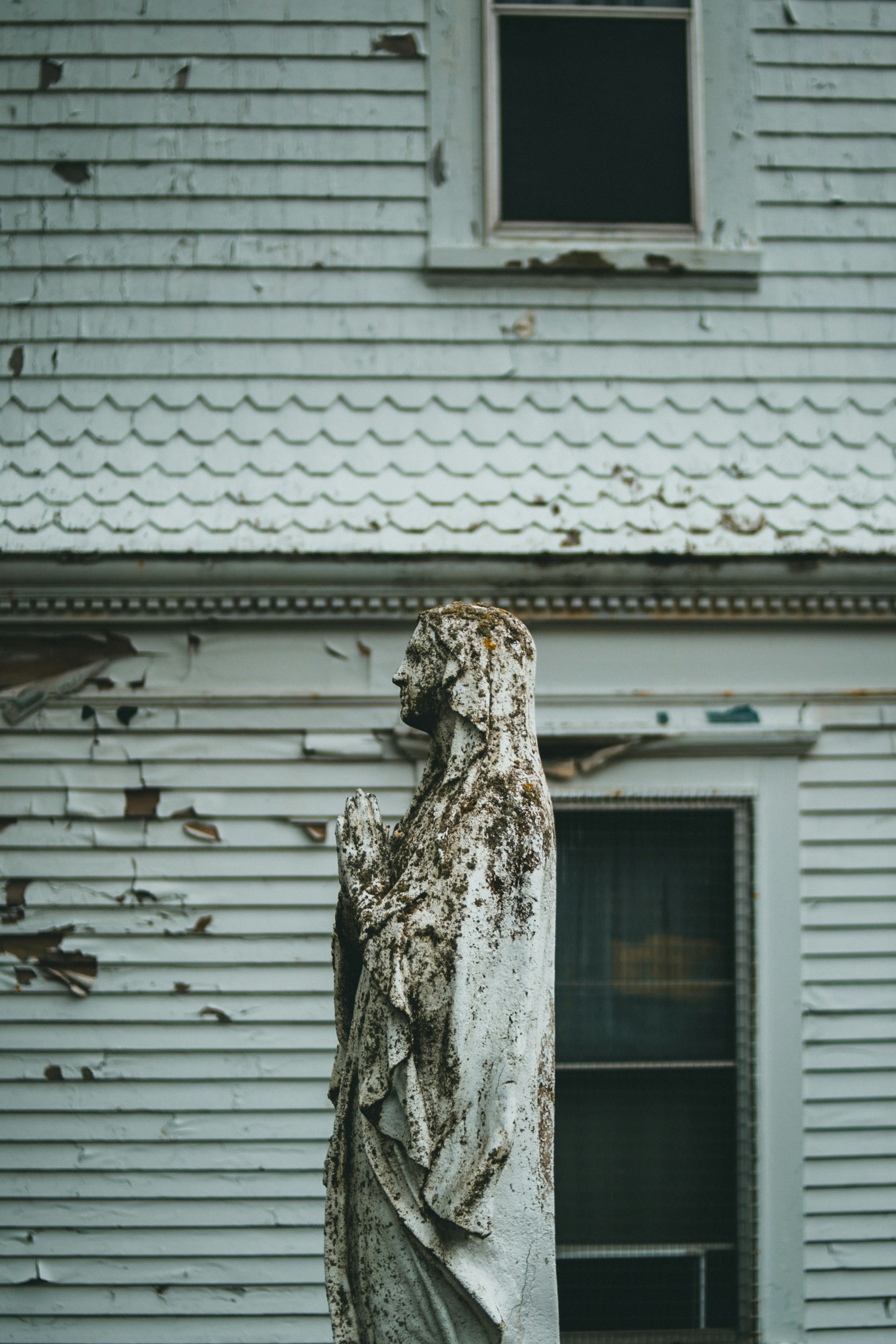 Weathered Sentinel: A Crumbling Statue Watches Over a Dilapidated House