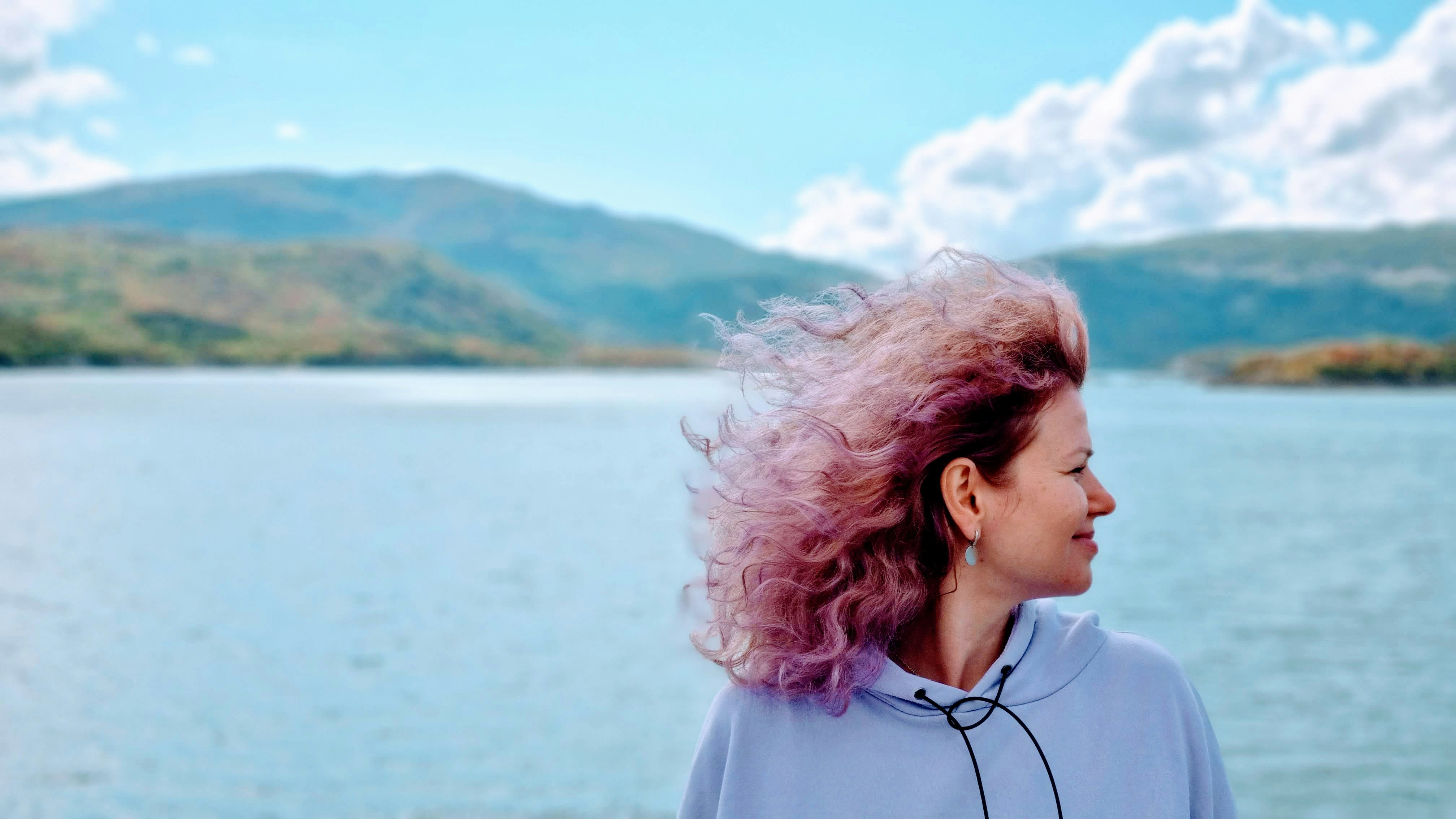 Woman with flowing pink hair gazes thoughtfully at a serene lake, framed by rolling hills and fluffy clouds.