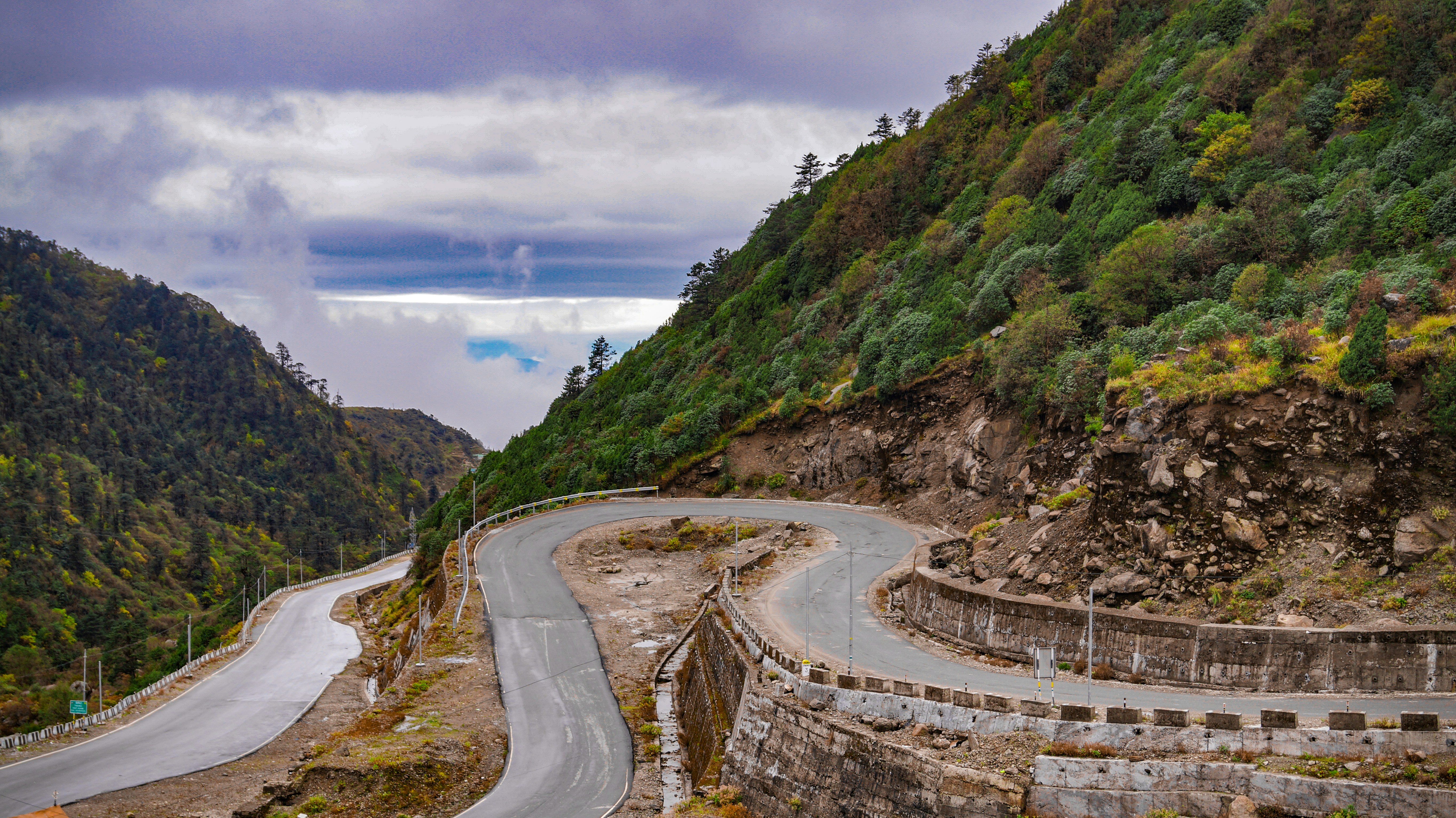 Curving road meanders through lush green hills under a cloudy sky, revealing a distant horizon. The scene captures the tranquil essence of mountainous terrain.