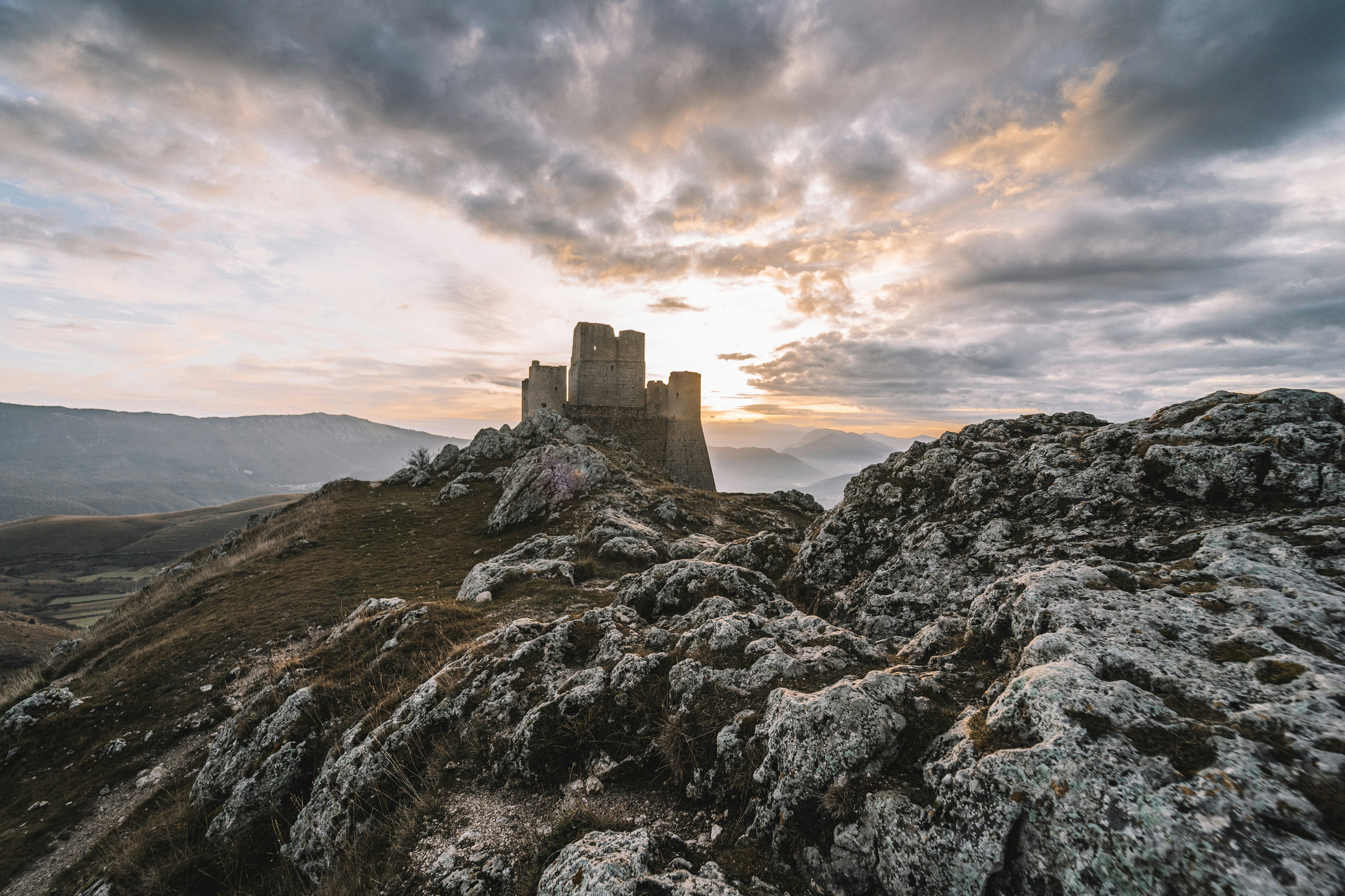 Ancient fortress perched atop rocky terrain under a dramatic sky at sunset. The scene captures the interplay of light and shadow across the landscape.