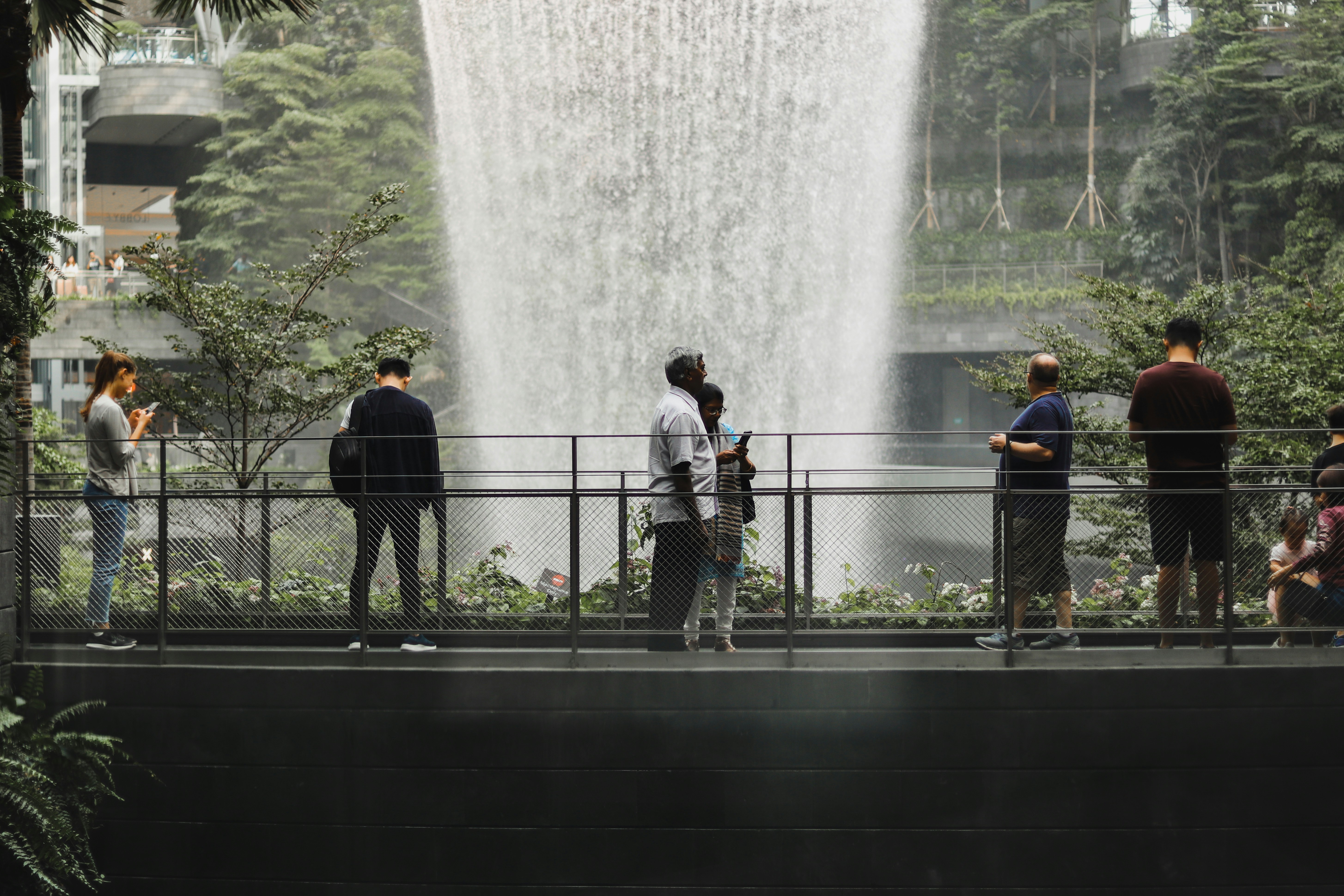 Visitors enjoy the tranquility of a waterfall while standing on a balcony surrounded by lush greenery. The scene captures a blend of nature and modern architecture.