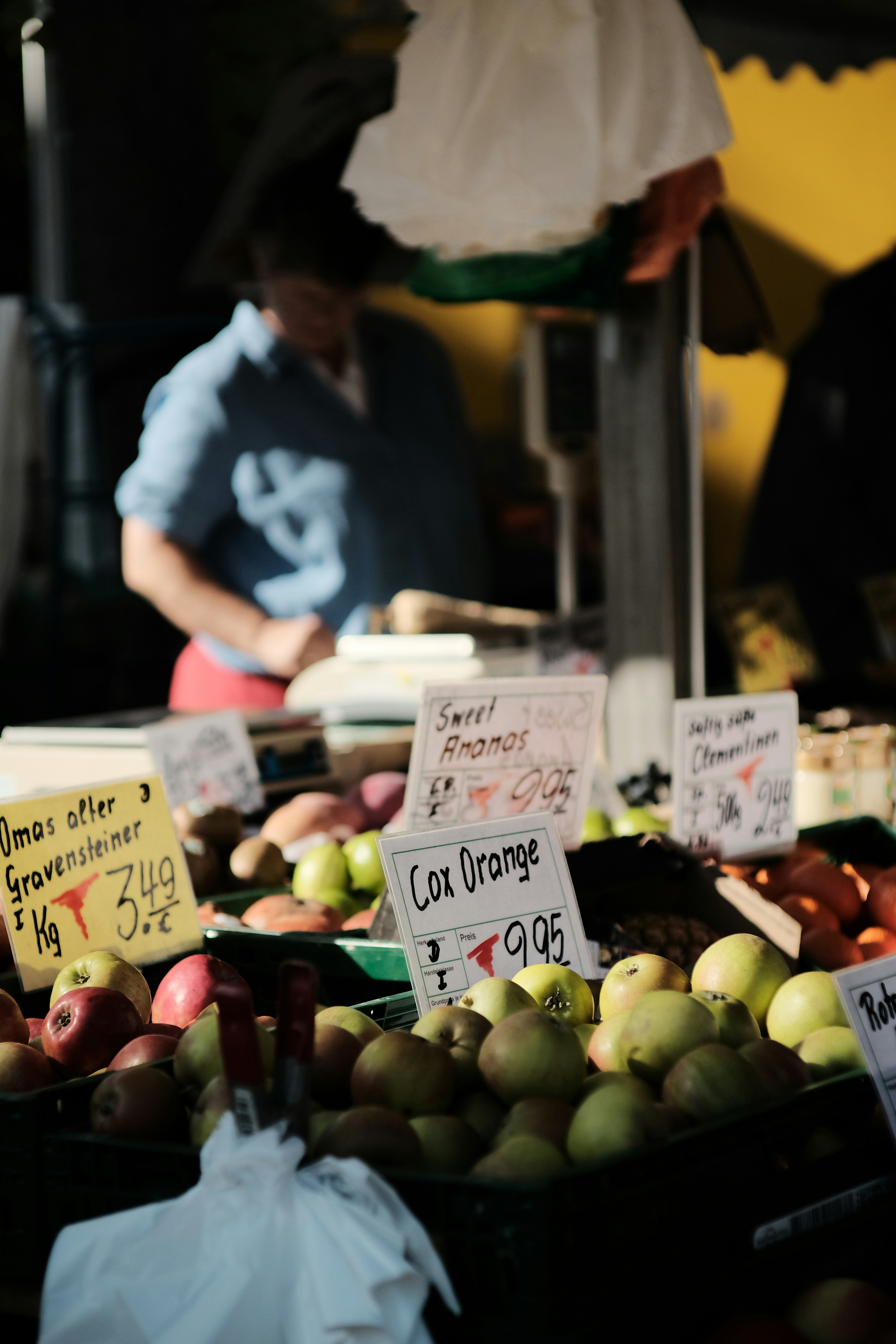 Vibrant assortment of fresh fruits displayed at a market stall, with price tags visible. A vendor is engaged in the background, adding life to the scene.