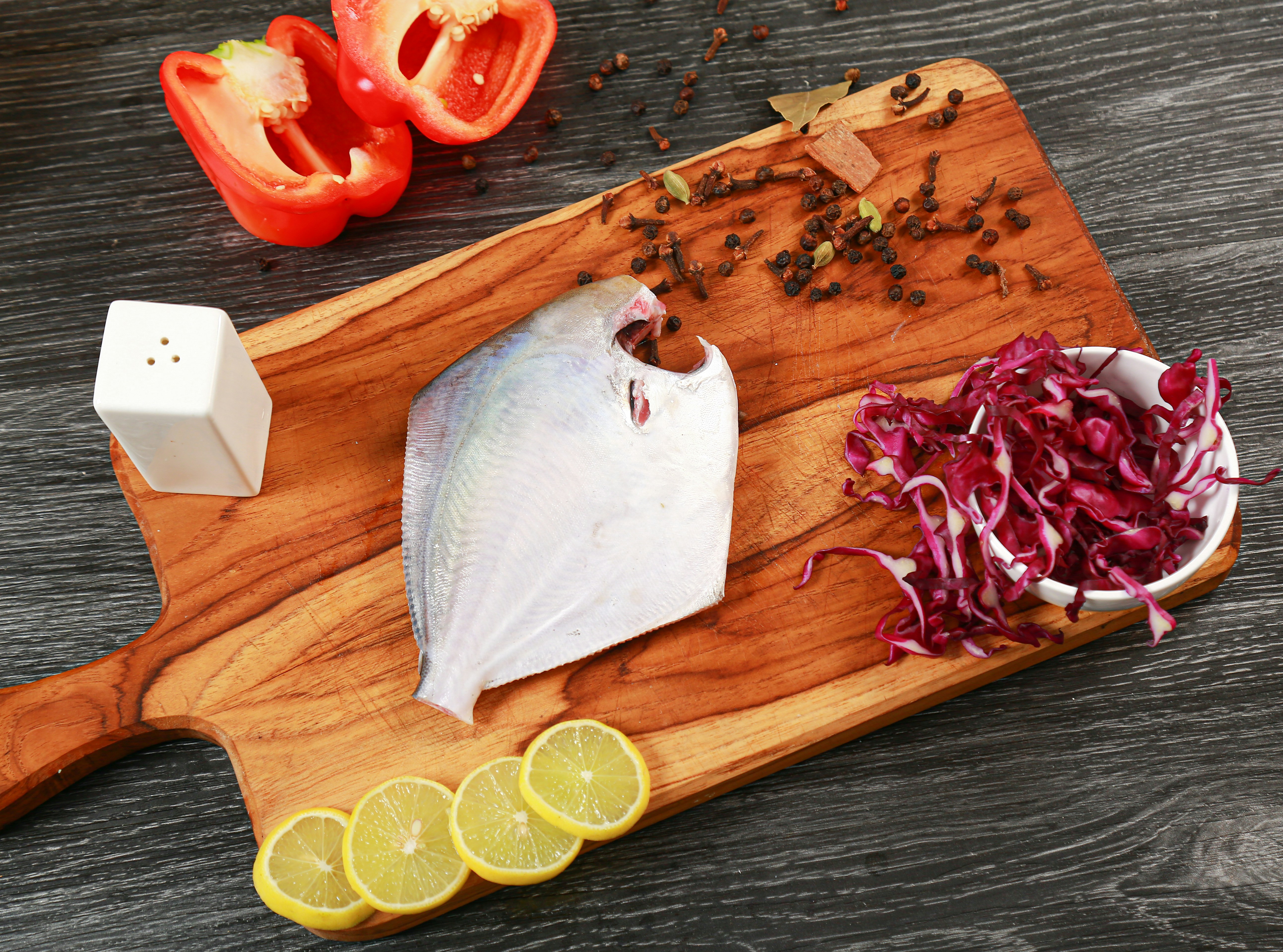 Fresh fish fillet accompanied by vibrant red bell peppers, red cabbage, spices, and lemon slices on a wooden cutting board.