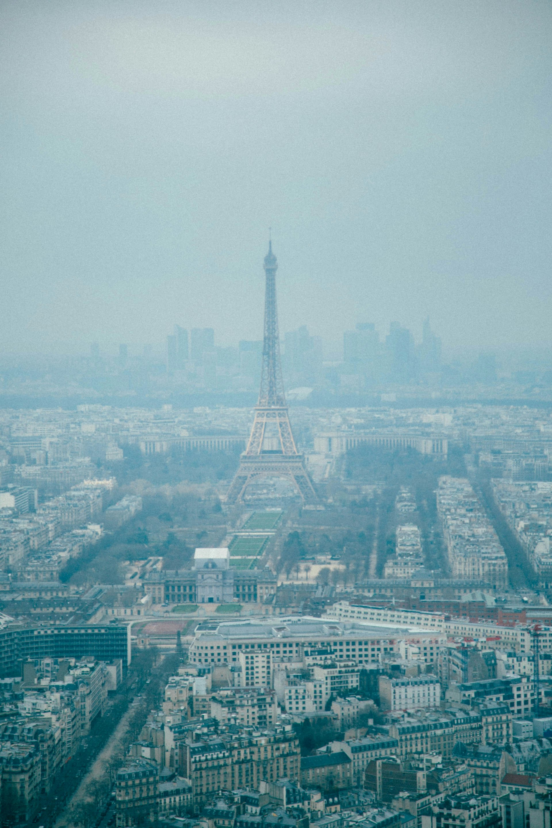 Eiffel Tower rising through a blanket of fog, surrounded by the sprawling cityscape of Paris. The scene captures the iconic structure's elegance amidst a muted atmosphere.