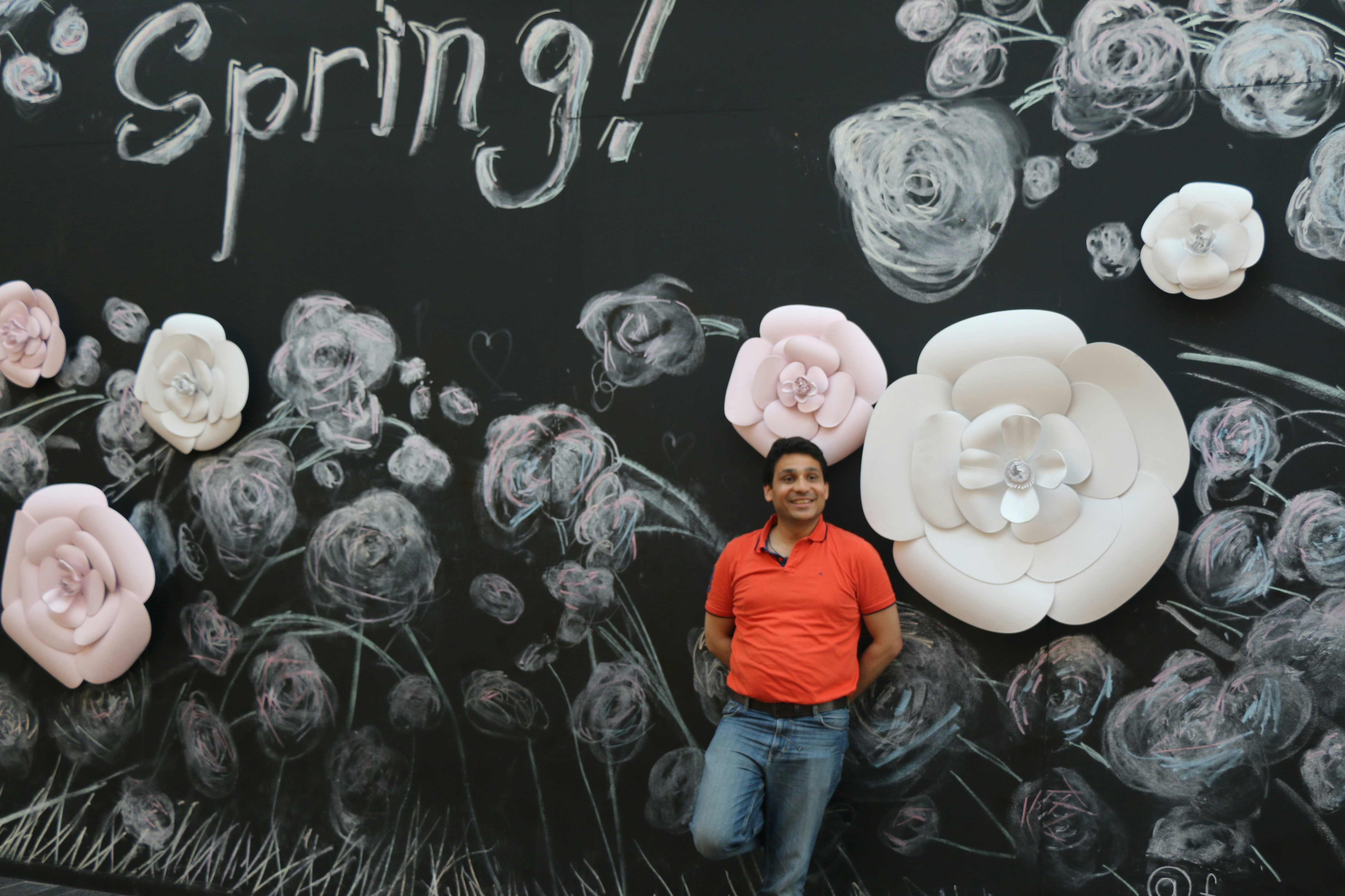 A cheerful young man poses against a vibrant mural adorned with large paper flowers and whimsical chalk drawings, celebrating the arrival of spring.