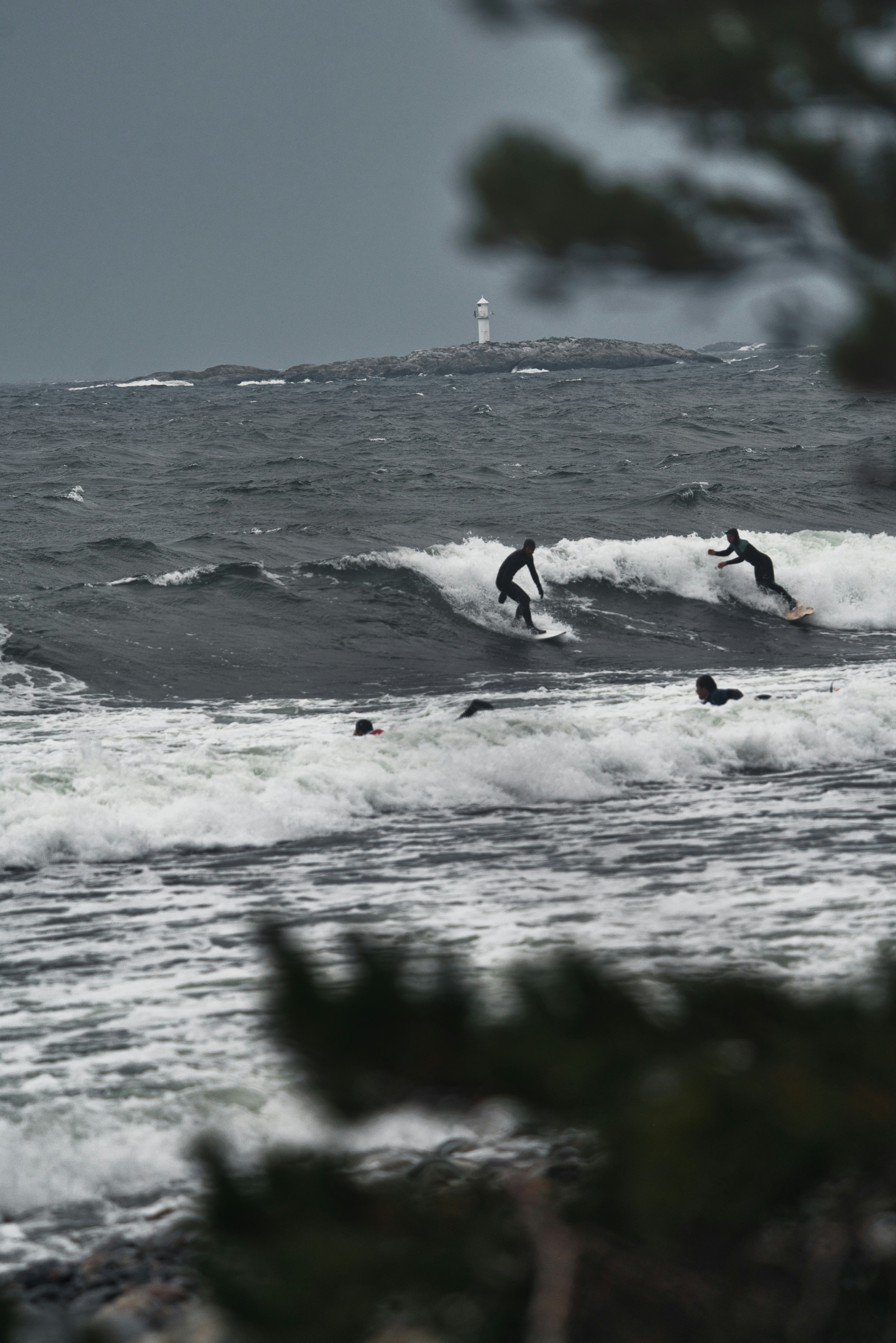Two surfers riding the waves under a moody sky, with a distant lighthouse adding context to the scene. The image conveys the thrill of surfing in challenging conditions.