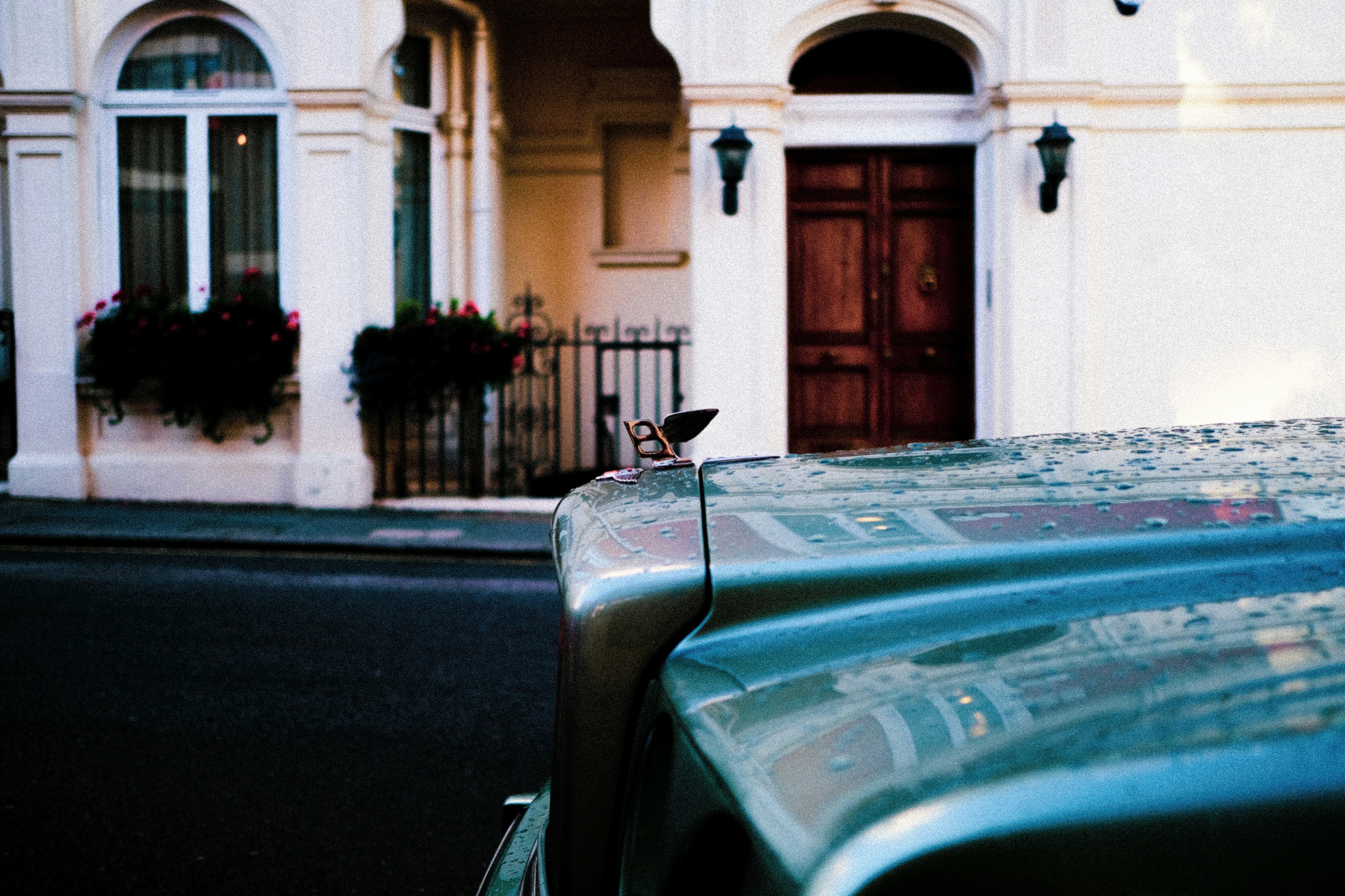 Vintage car hood with a decorative ornament, set against a charming backdrop of classic architecture and blooming flowers.
