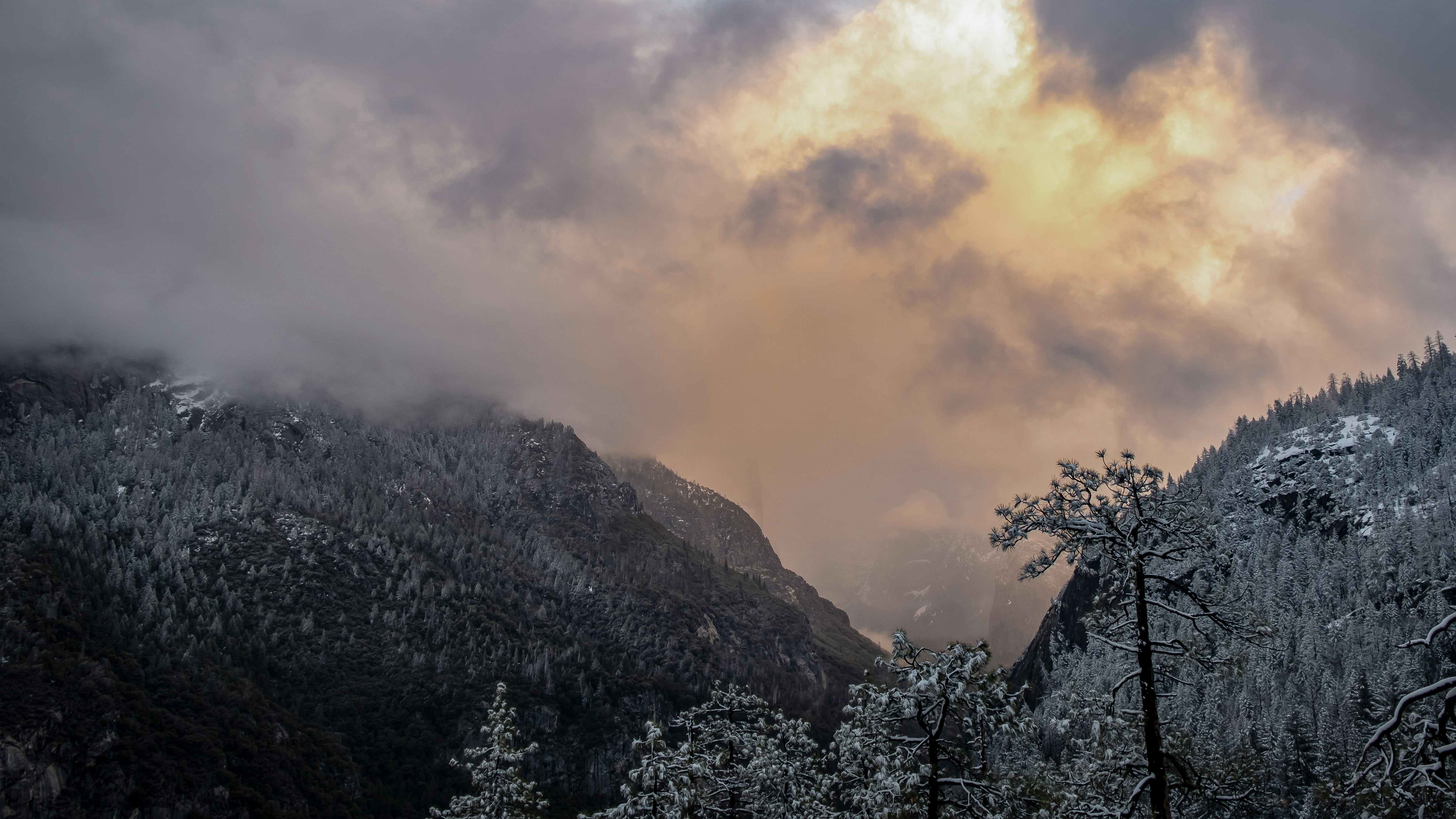 Snow-dusted mountains loom under a dramatic sky, where clouds part to reveal a warm glow at dawn.