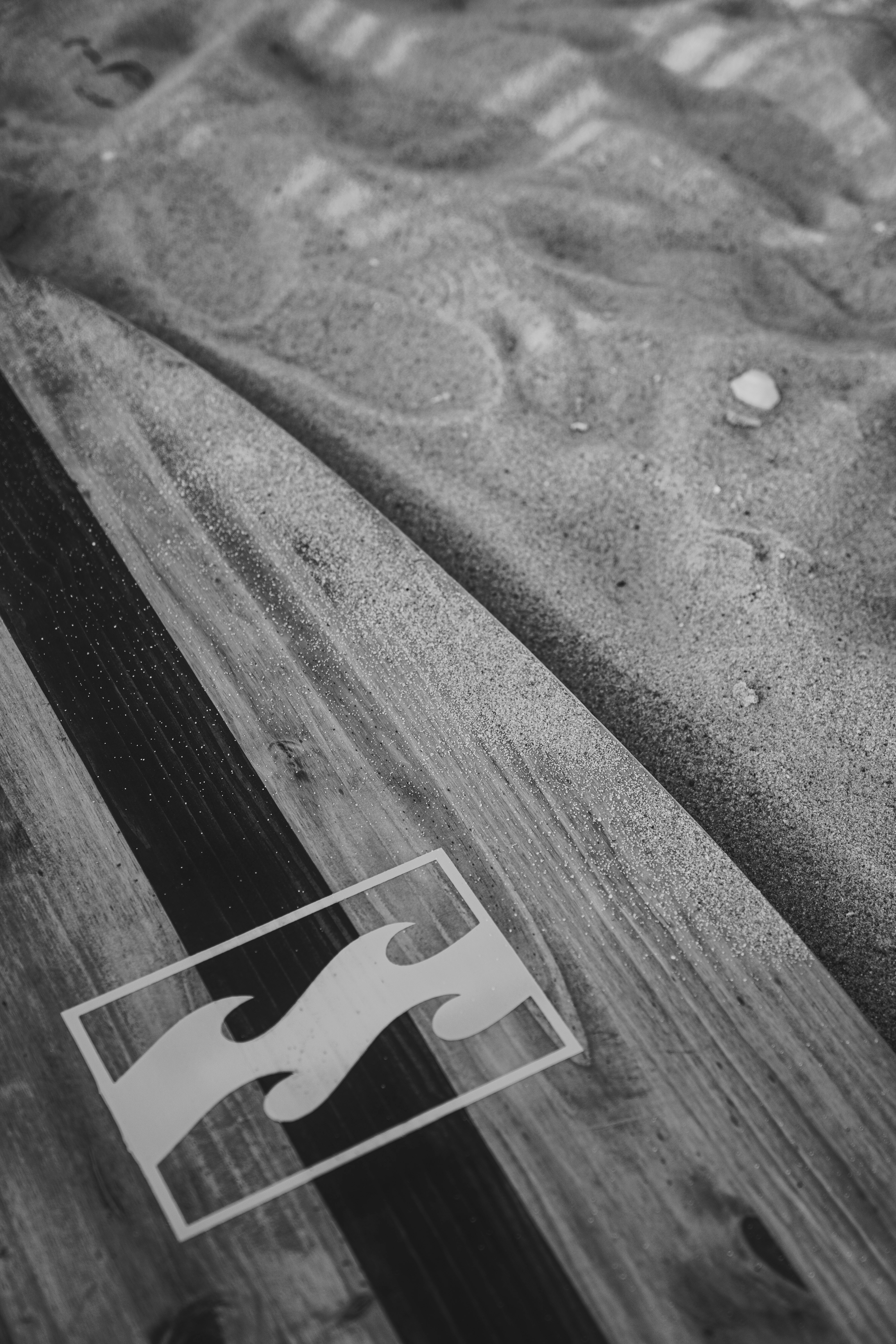Close-up of a surfboard resting on sandy beach, showcasing its logo and wood grain details. The monochrome tones emphasize the textures and natural elements.