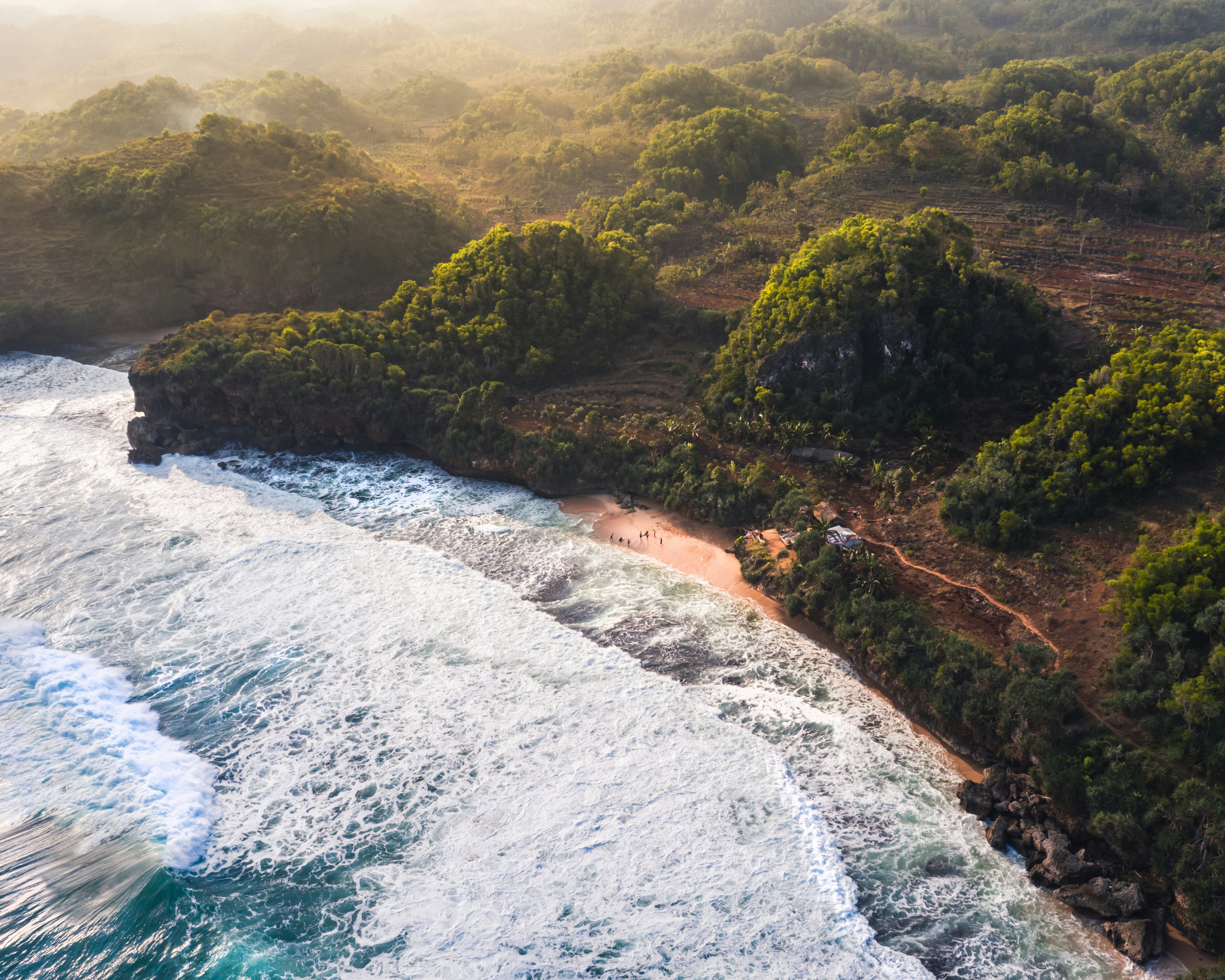 Aerial view of ocean waves crashing against a rocky, forested coastline under a hazy sky.