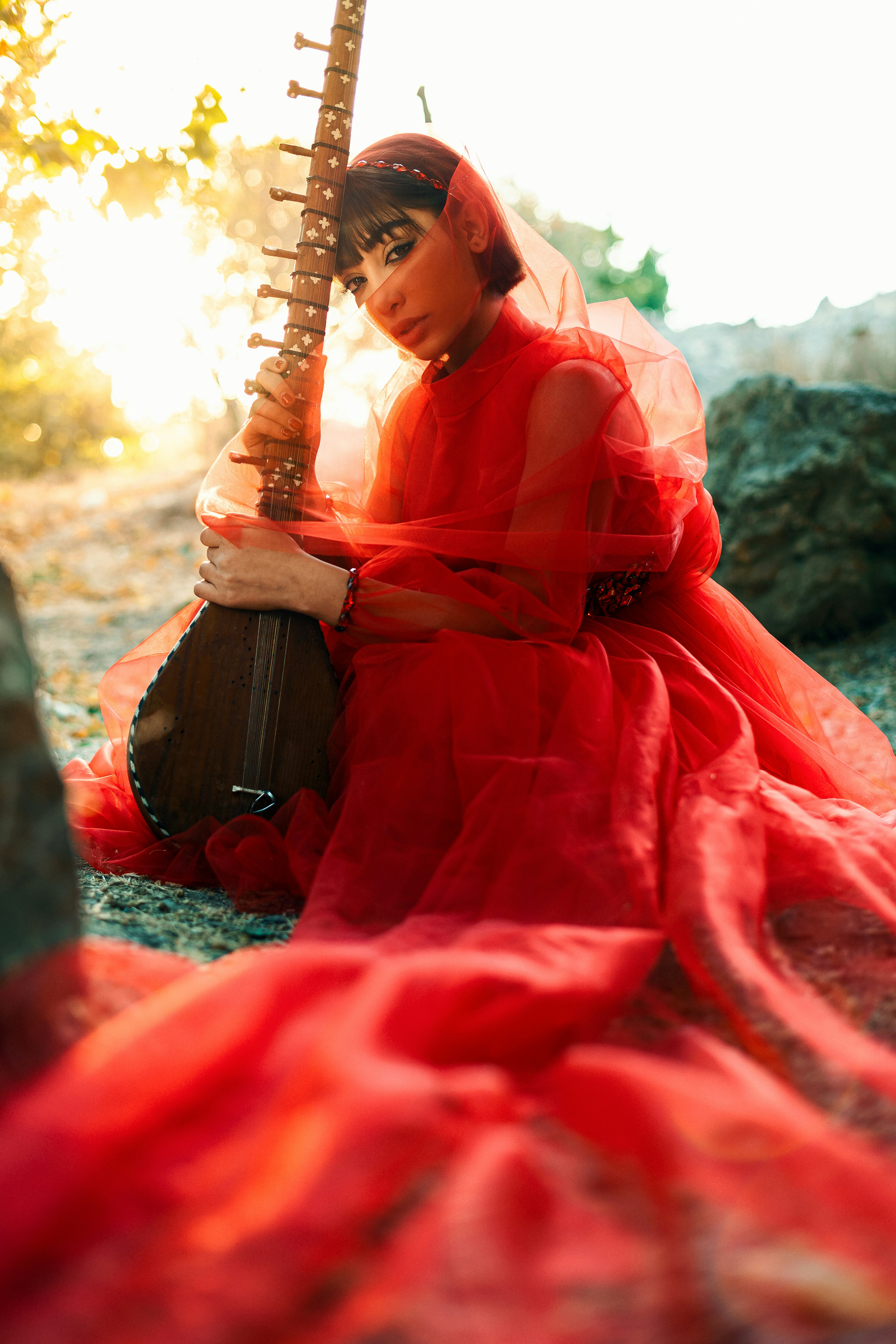 Woman in a flowing red dress holds a traditional string instrument, surrounded by soft sunlight filtering through trees. The scene evokes a sense of harmony and cultural depth.