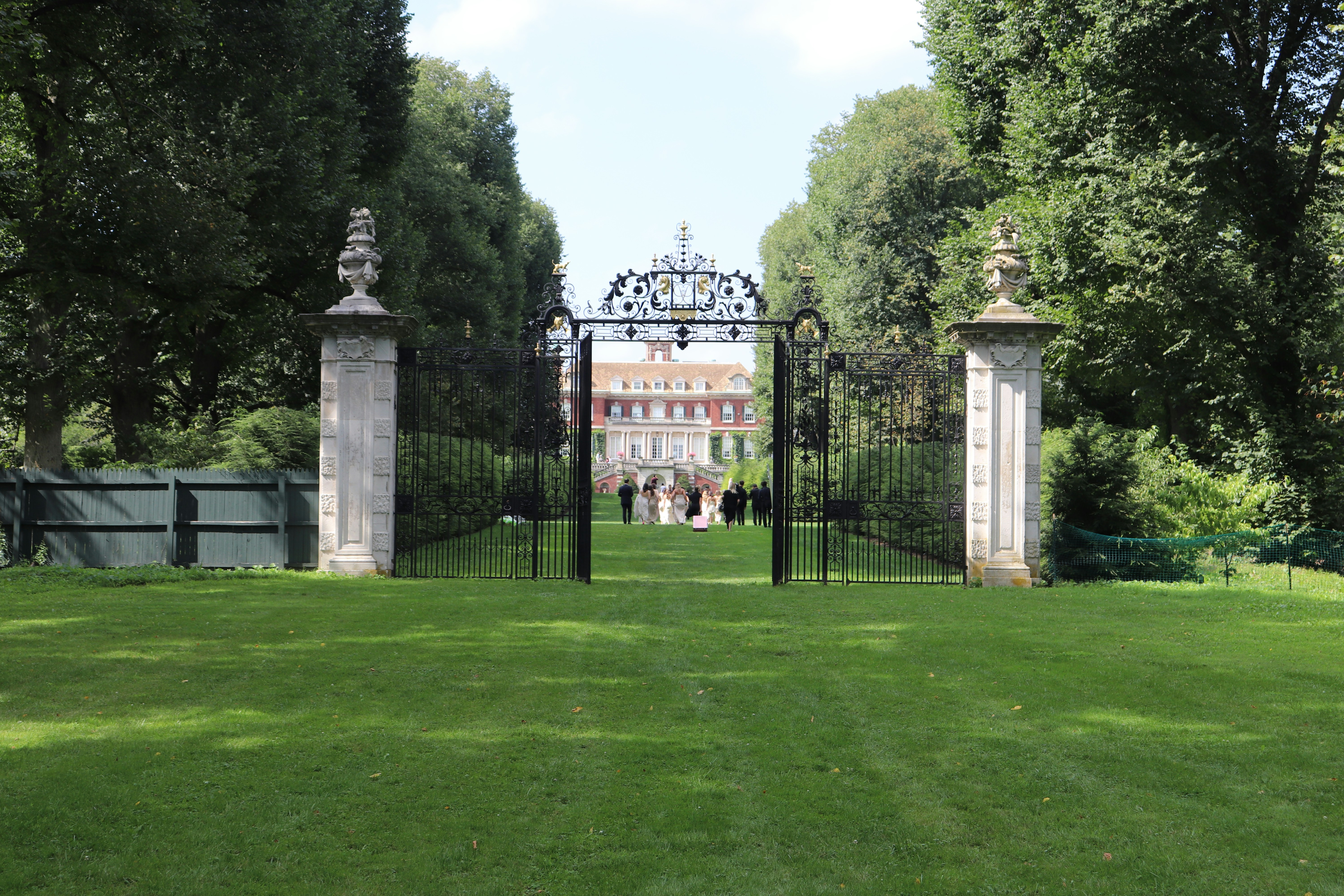 Intricate wrought iron gates open to a manicured lawn leading to a grand estate, framed by lush greenery.