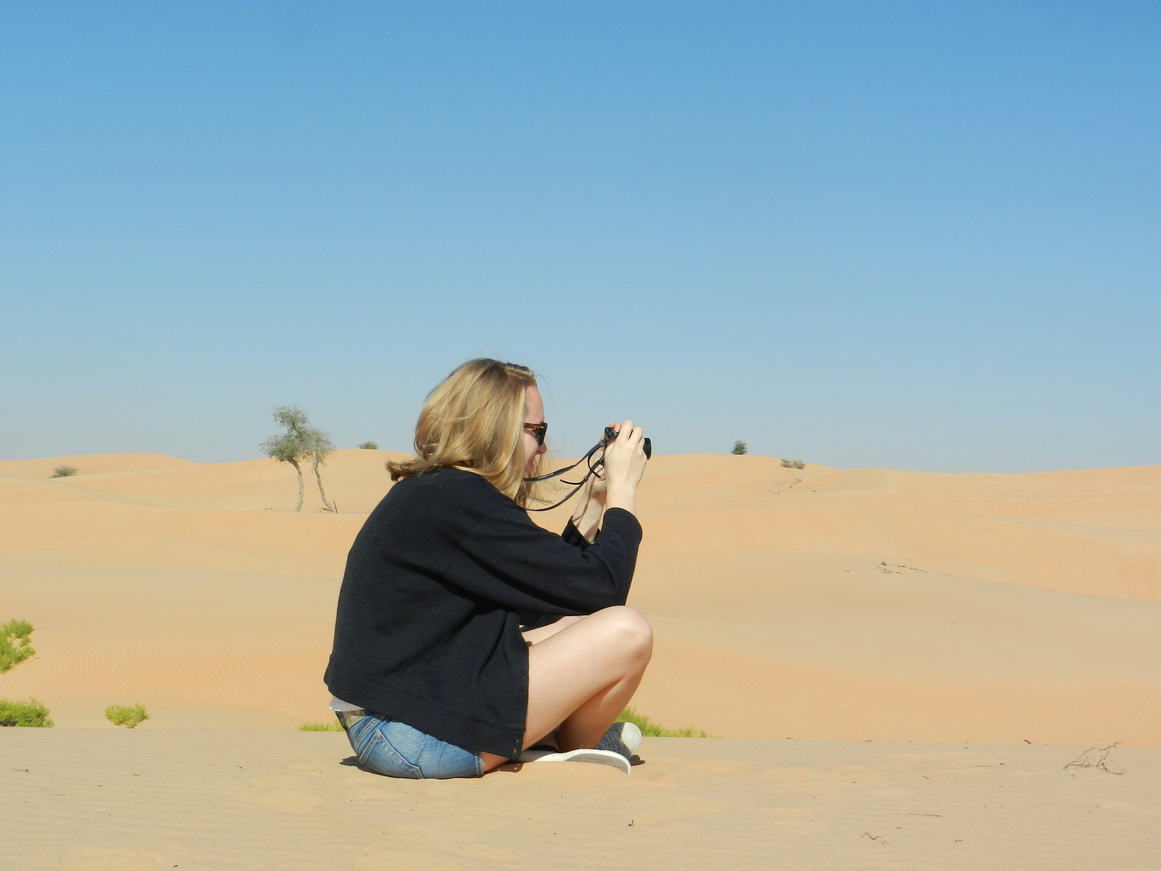 A woman crouches in the desert, focused on photographing the vast sandy landscape. The clear blue sky contrasts with the warm tones of the dunes.