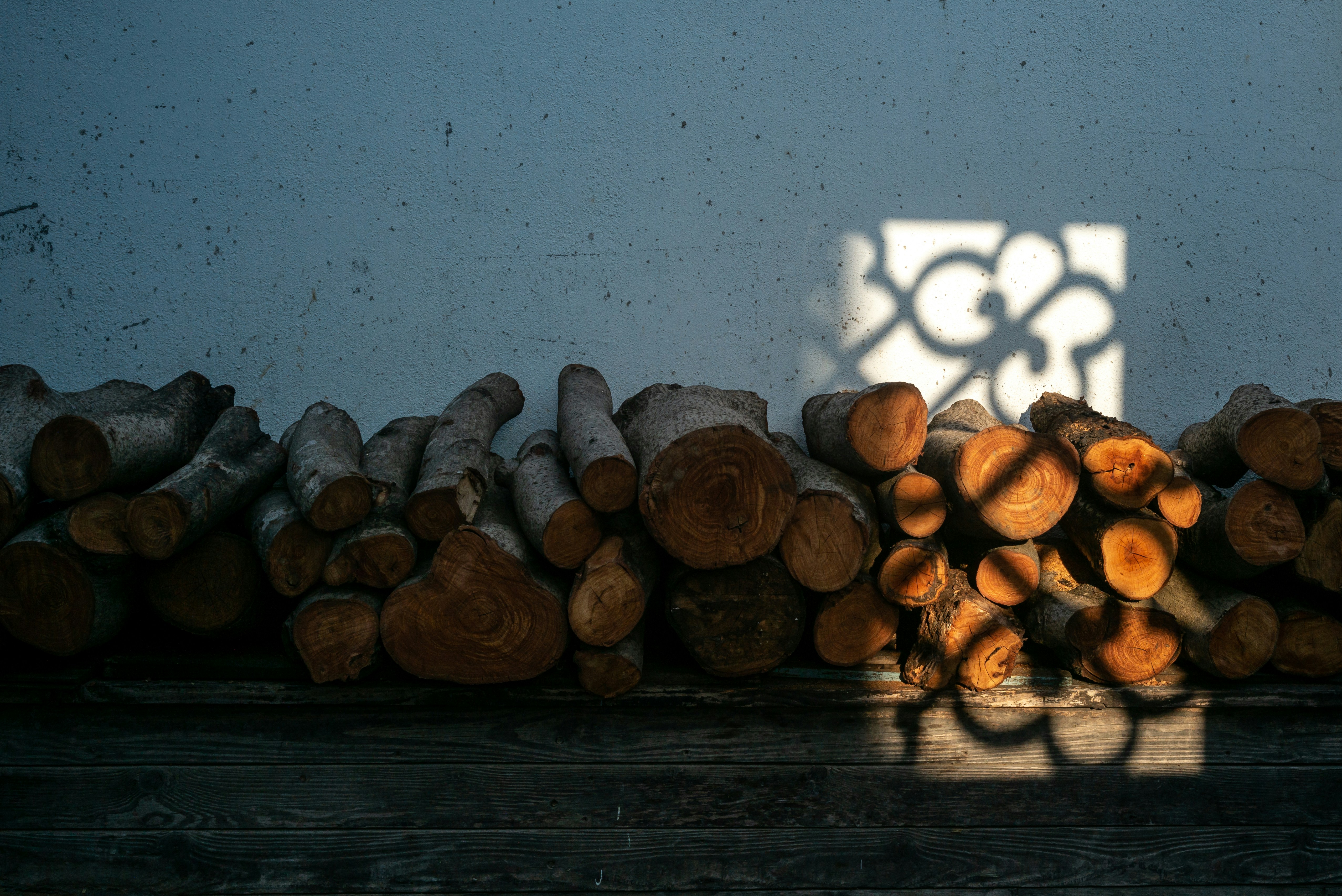 Still-life photograph of a stack of chopped logs on a dark wooden surface, bathed in warm light. A geometric lattice shadow on the gray wall adds contrast.
