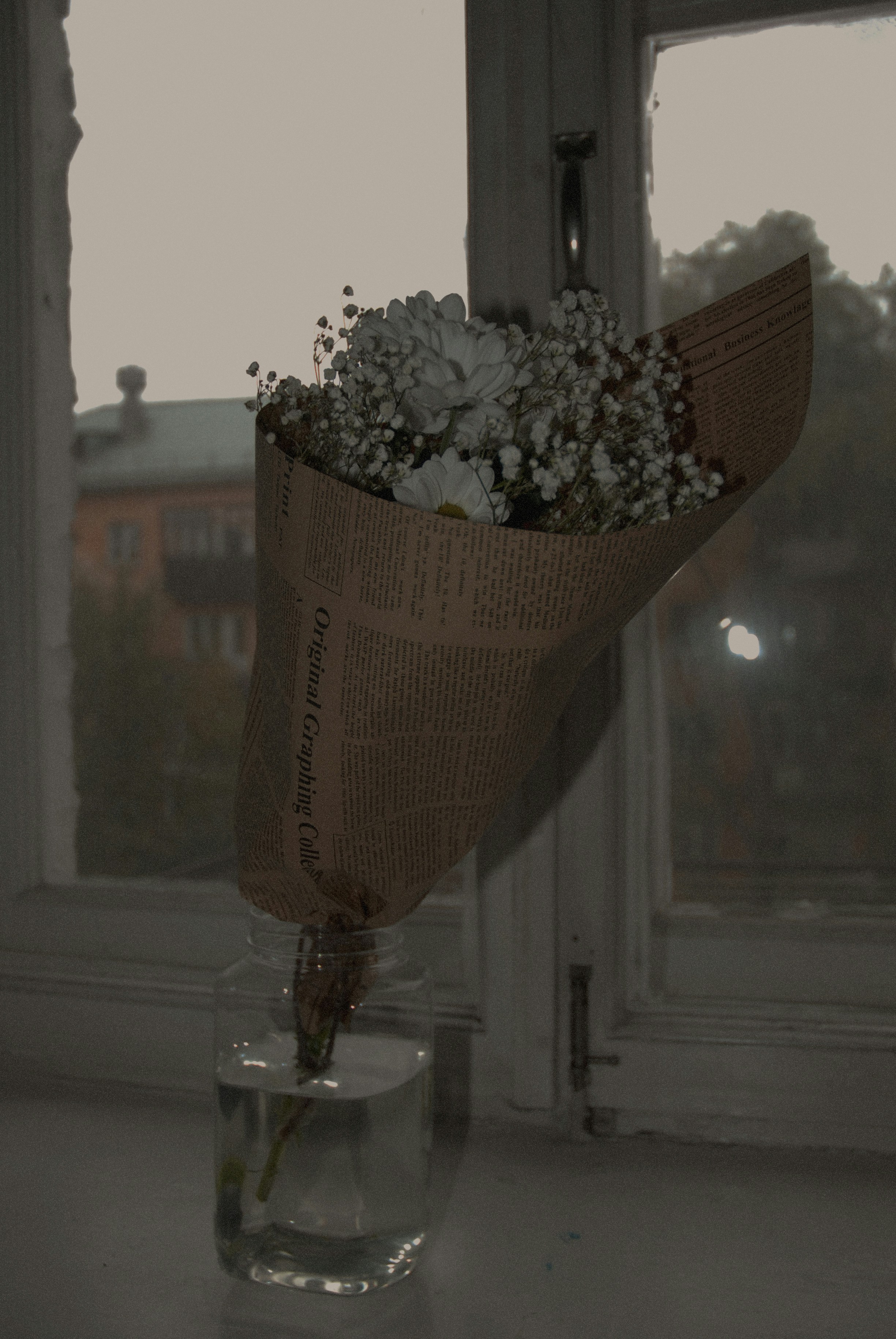 A bouquet of white flowers wrapped in newspaper rests in a glass jar filled with water, positioned near a window with a soft, muted background.