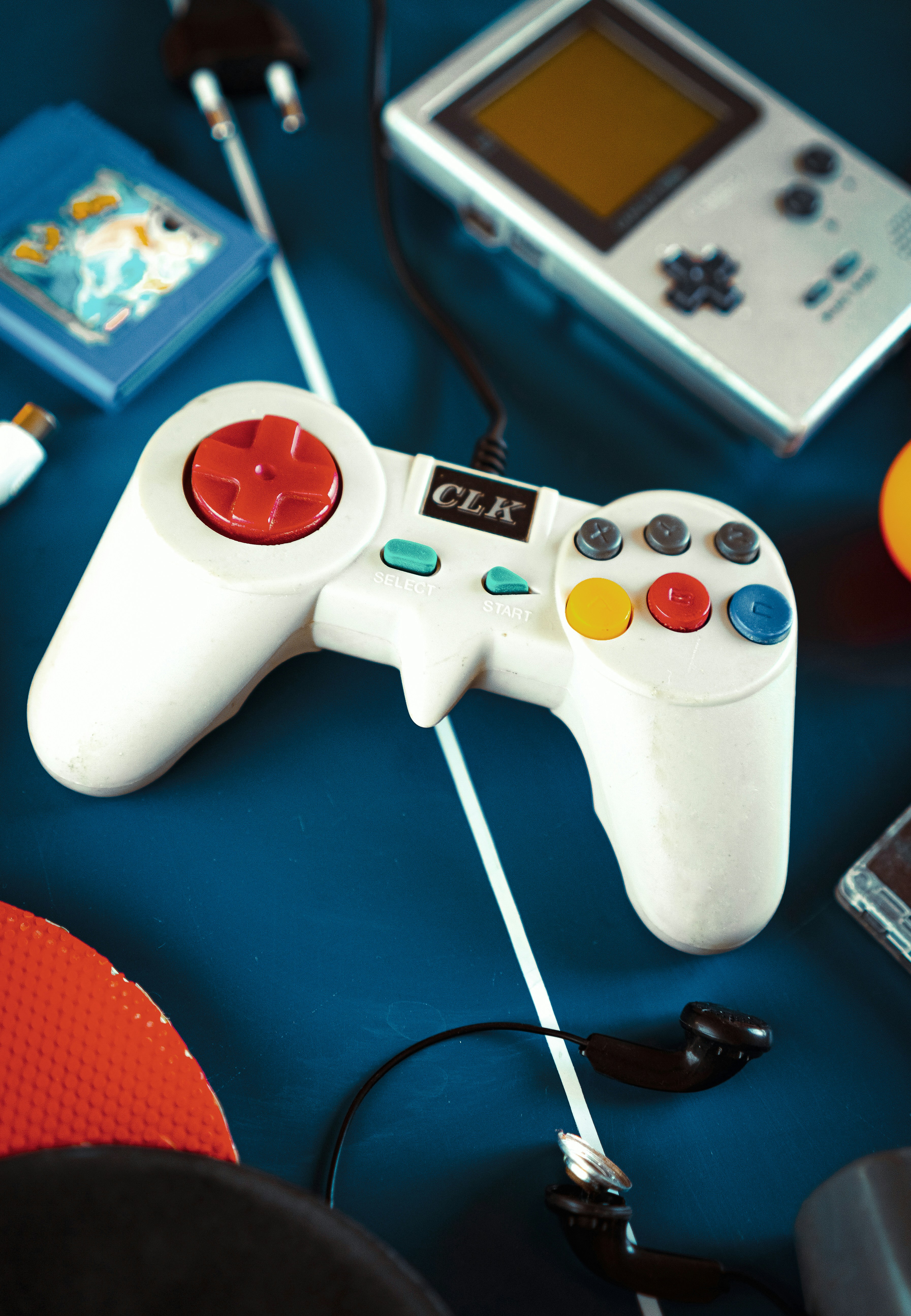 Colorful game controller sits prominently on a blue surface among vintage gadgets; a handheld console and wires frame the scene, suggesting a playful tech setup.