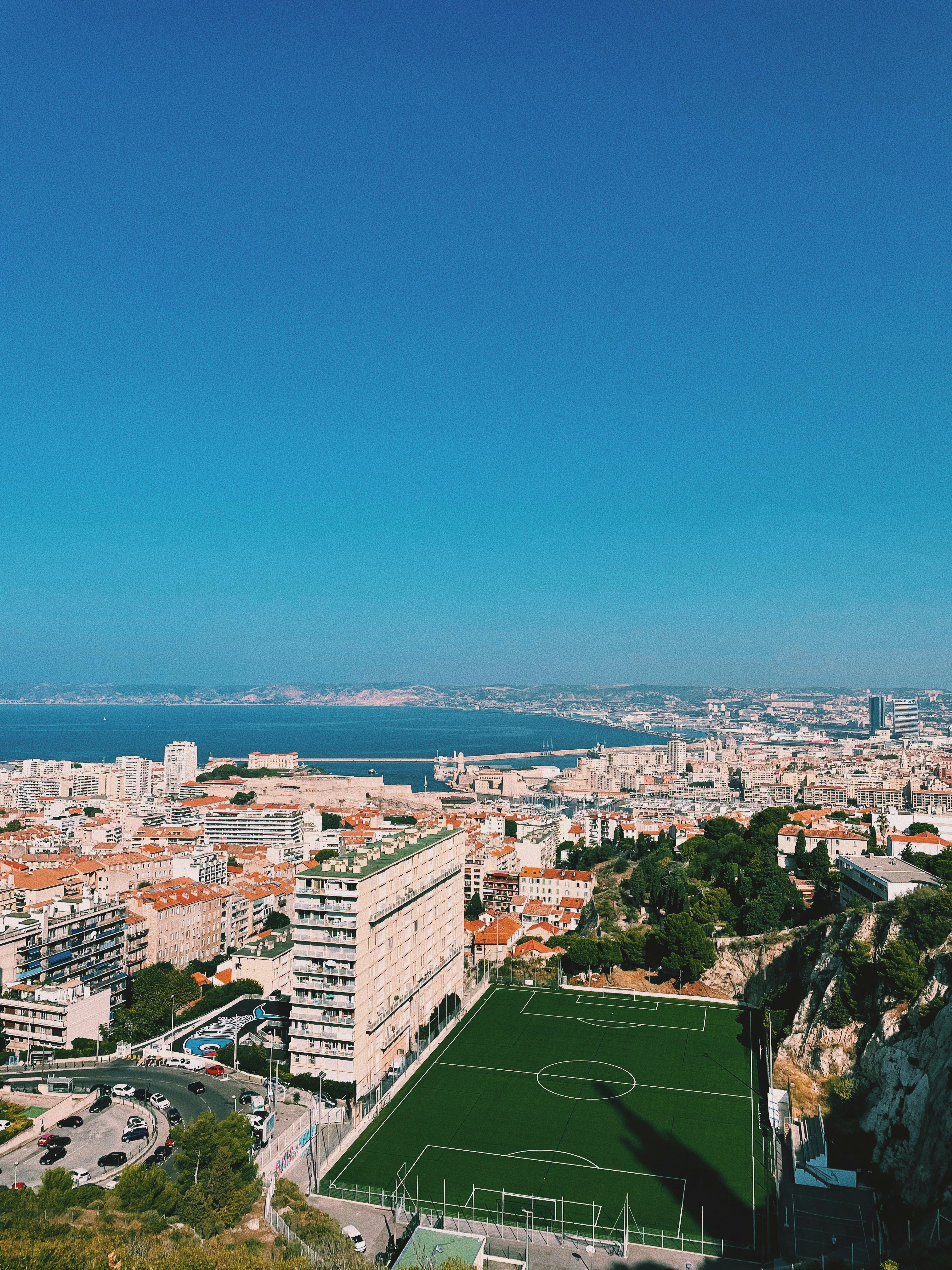 A panoramic view of a coastal city featuring a vibrant green soccer field, residential buildings, and the shimmering sea in the background.