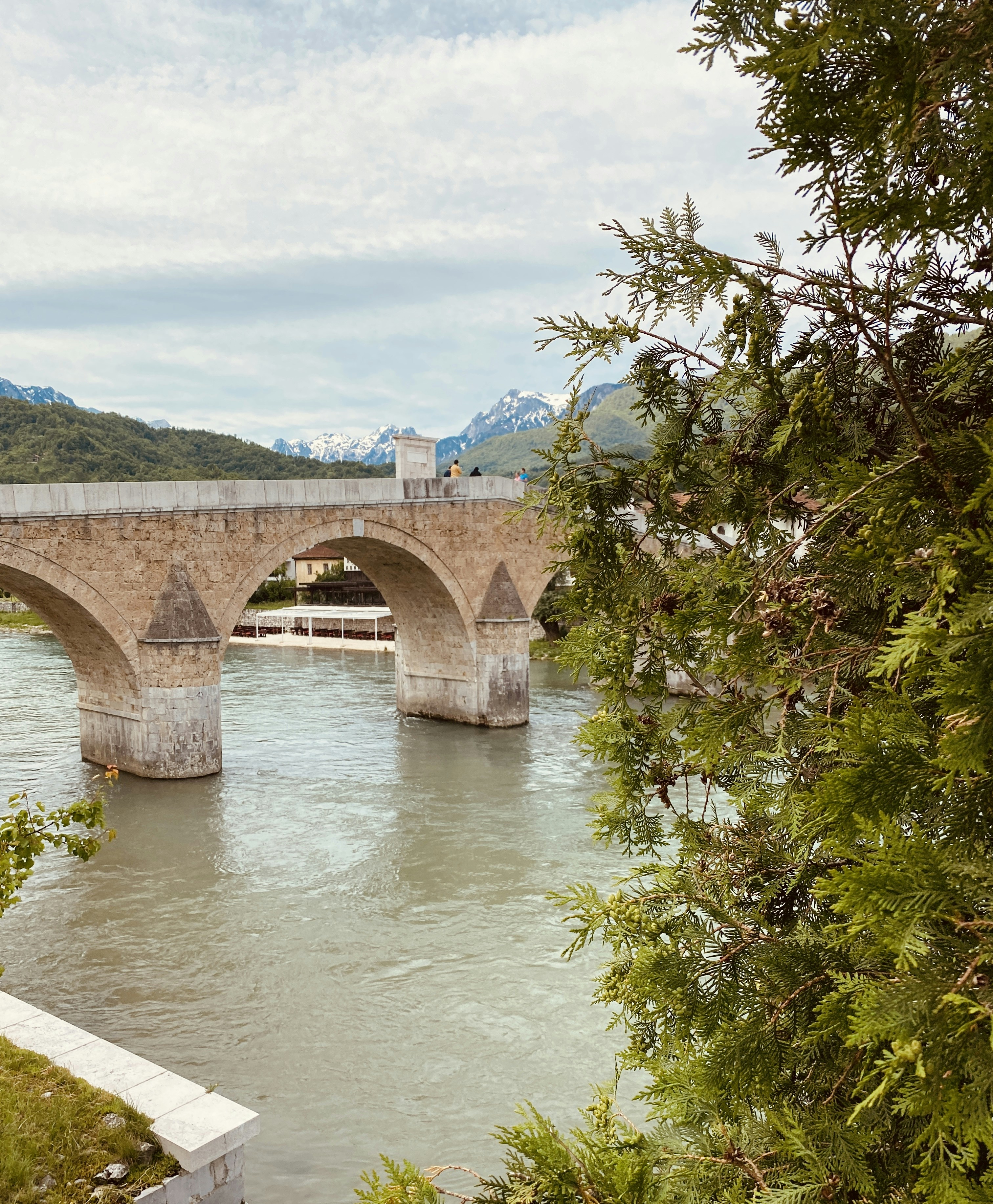 Historic stone bridge arching over a serene river, framed by lush greenery and distant snow-capped mountains.