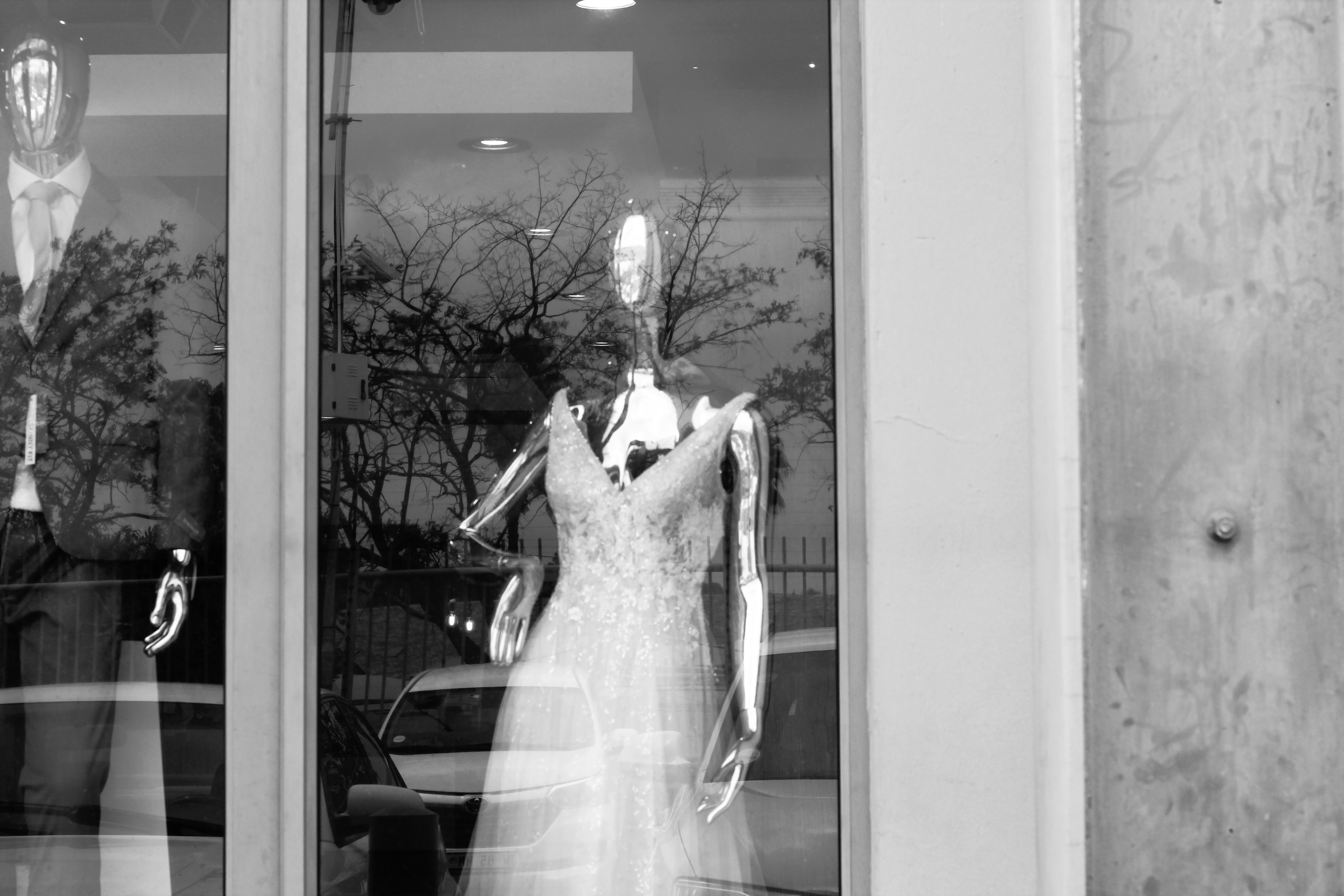 Mannequins in a shop window display elegant clothing with reflections of trees and cars outside.