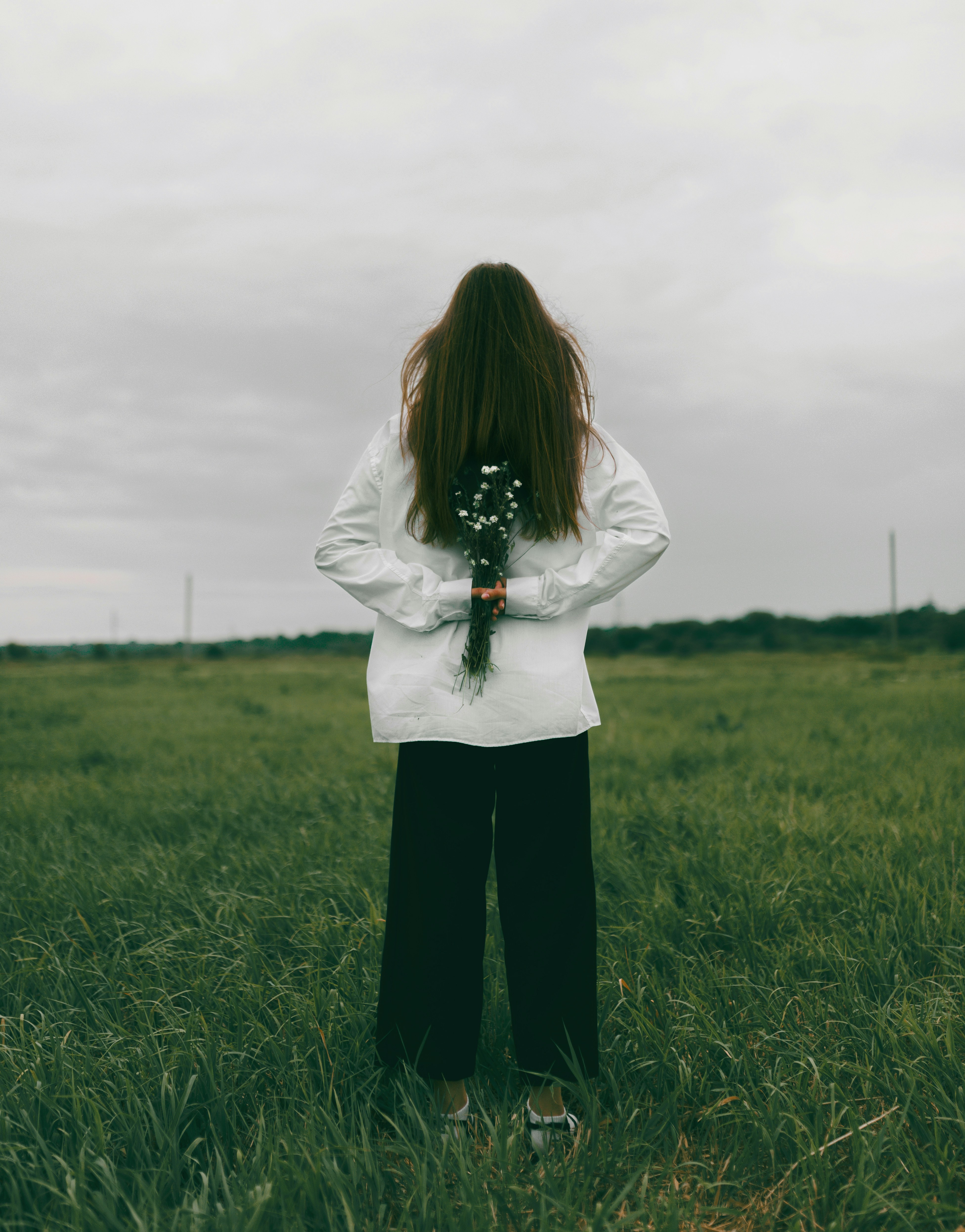 Person standing in a grassy field, holding a bouquet of wildflowers behind their back, with a soft, overcast sky above.