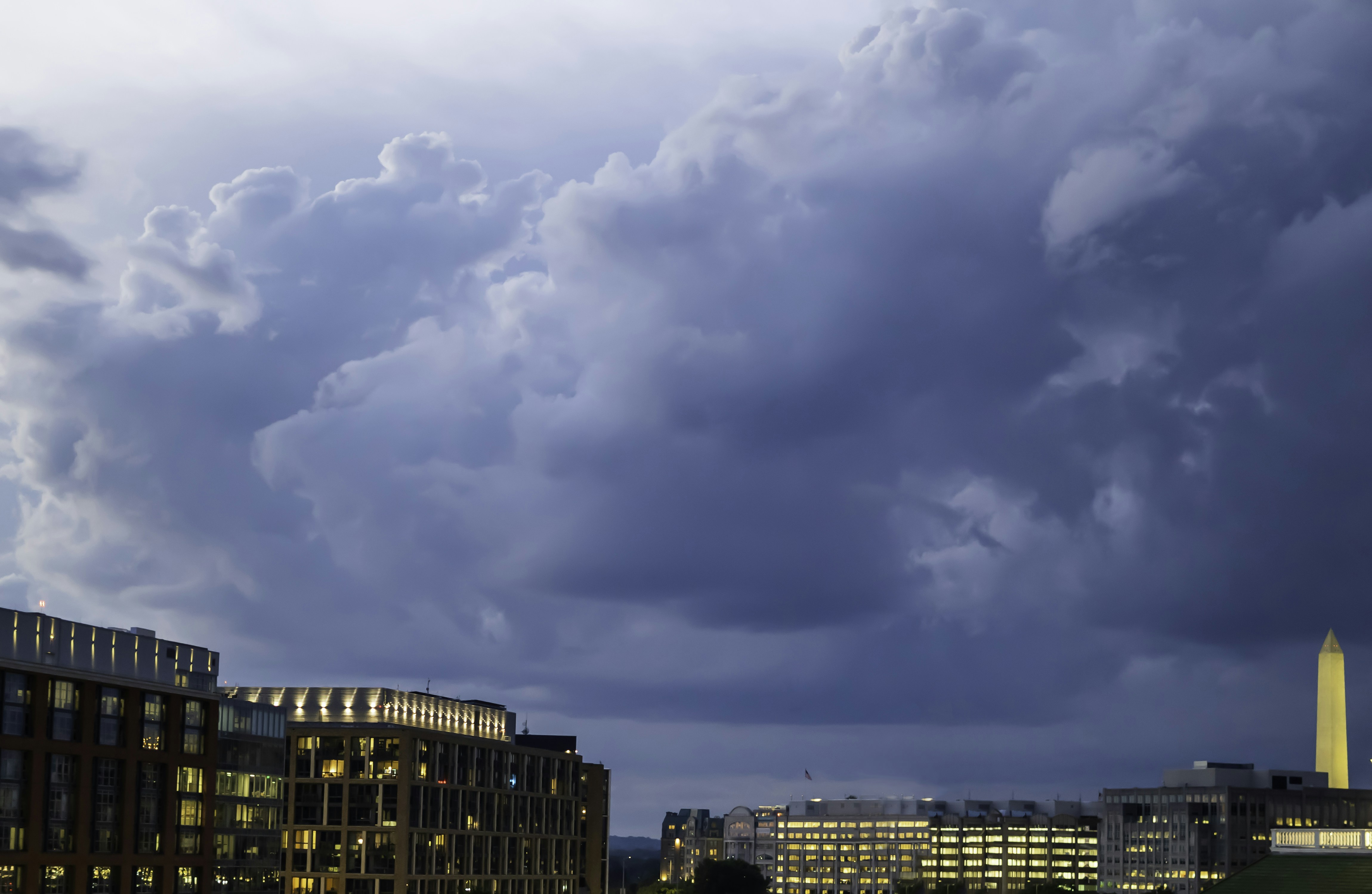Dramatic clouds loom over a city skyline, highlighting the contrast between the illuminated buildings and the darkening sky. The Washington Monument stands tall in the distance.