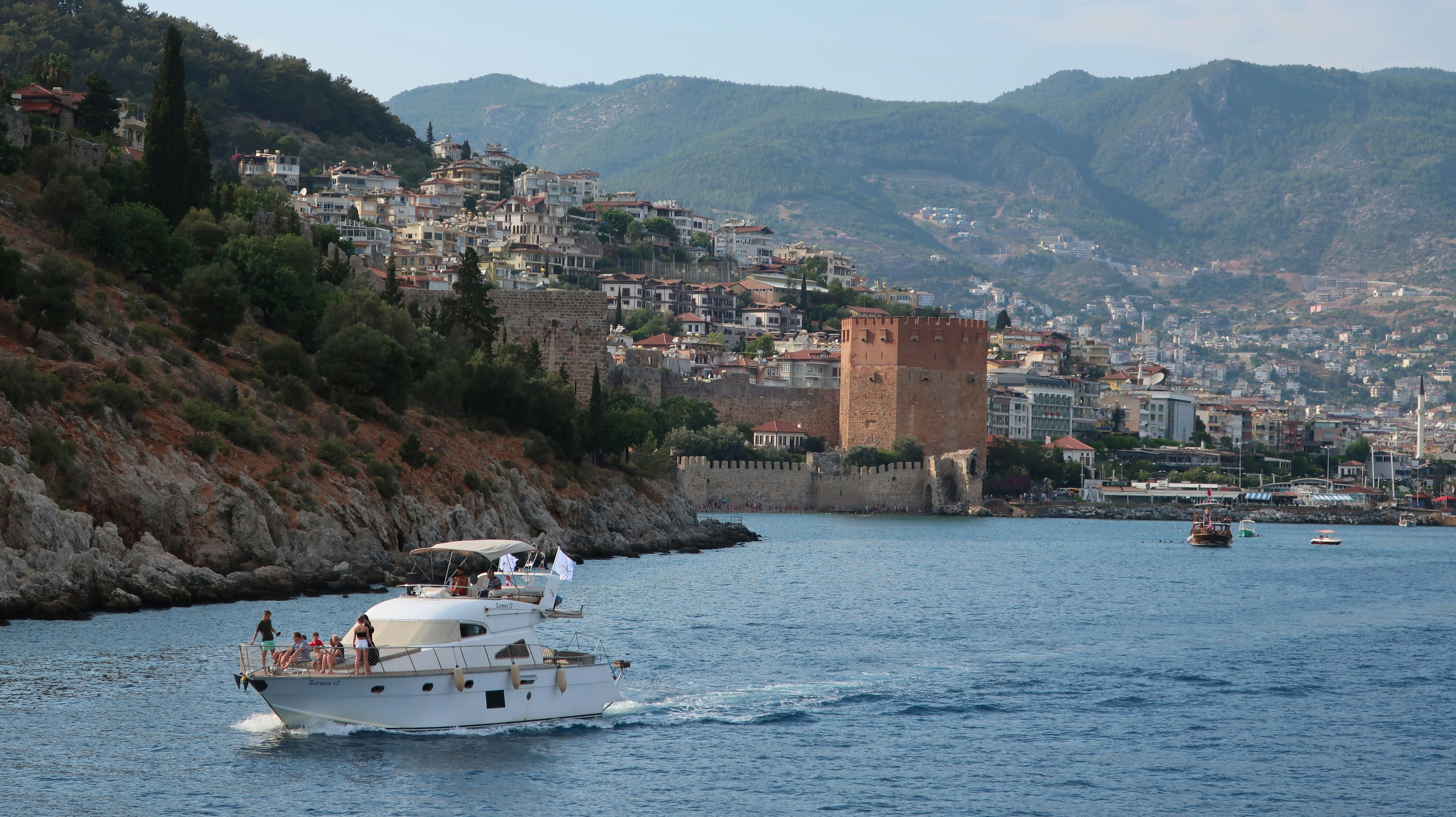 White yacht cruising along a coastal town with mountainous backdrop under clear skies.
