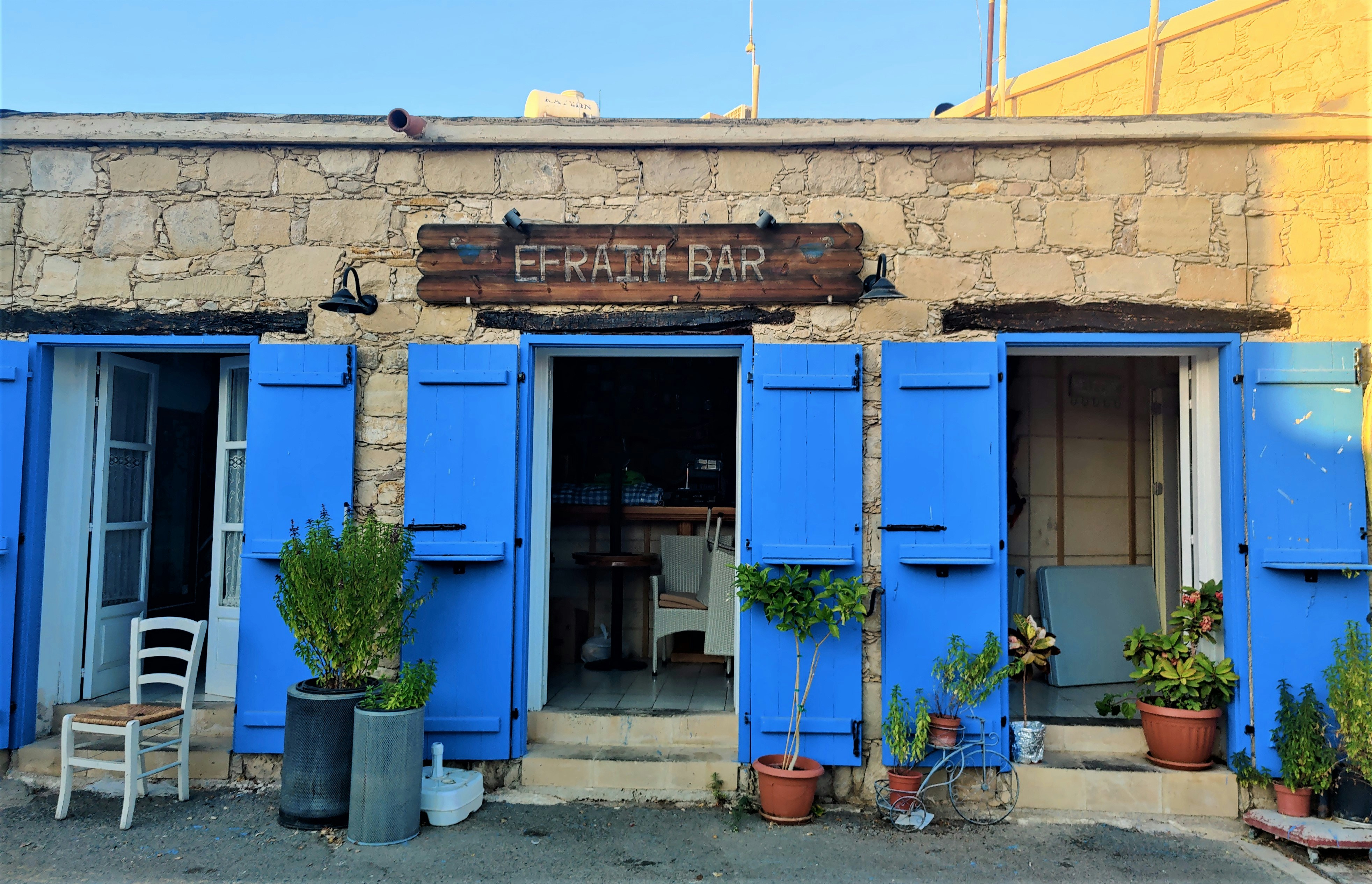 Quaint bar entrance with vibrant blue shutters and a rustic stone facade, surrounded by potted plants. The inviting atmosphere suggests a perfect spot for relaxation.