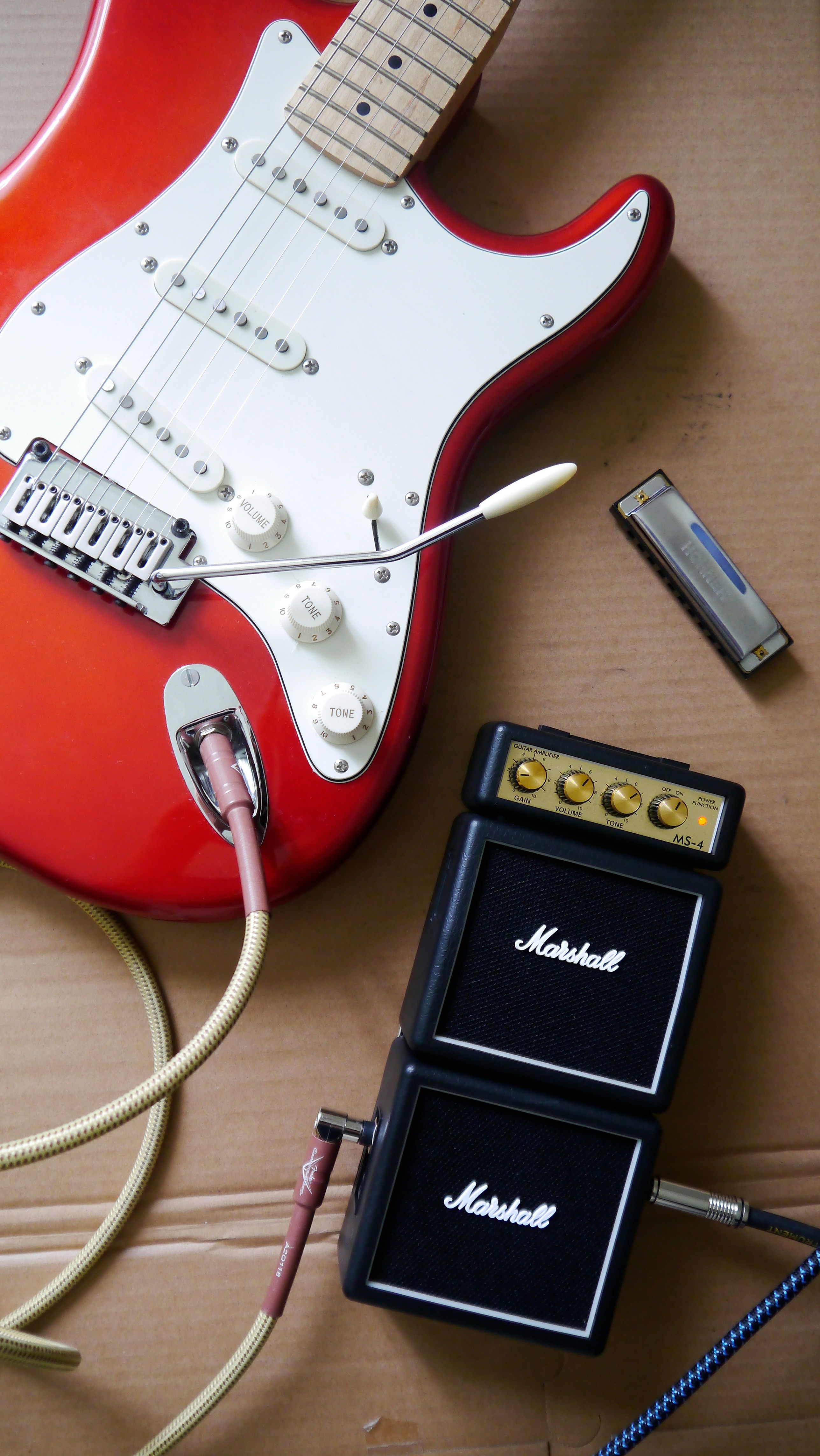 An electric guitar with a vibrant red finish alongside a Marshall amplifier and harmonica, arranged on a cardboard surface.